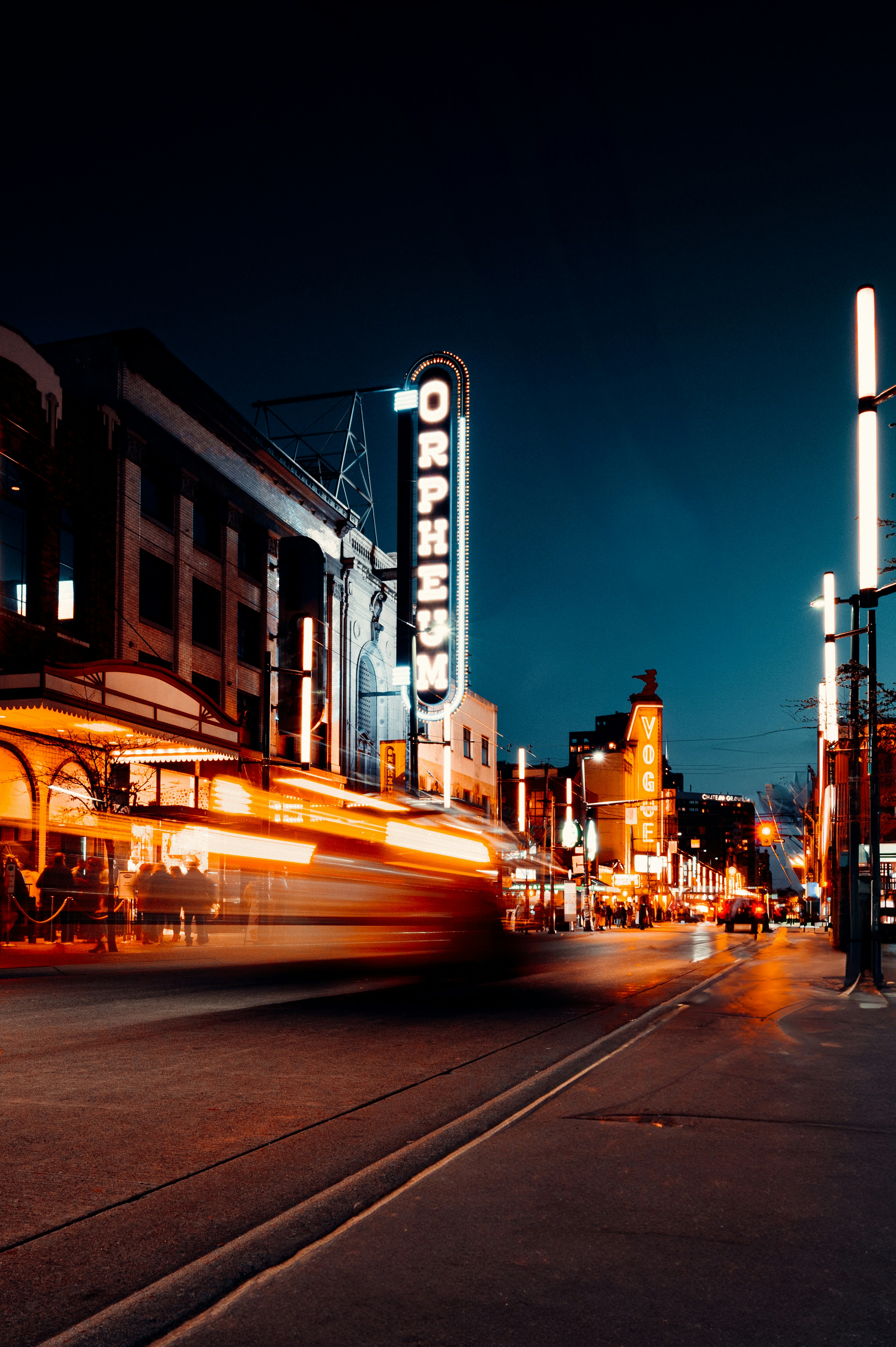Neon theater signs illuminate a vintage downtown street as traffic creates red-orange light trails. A long-exposure photograph captures a moody blue night sky and warm street glow.