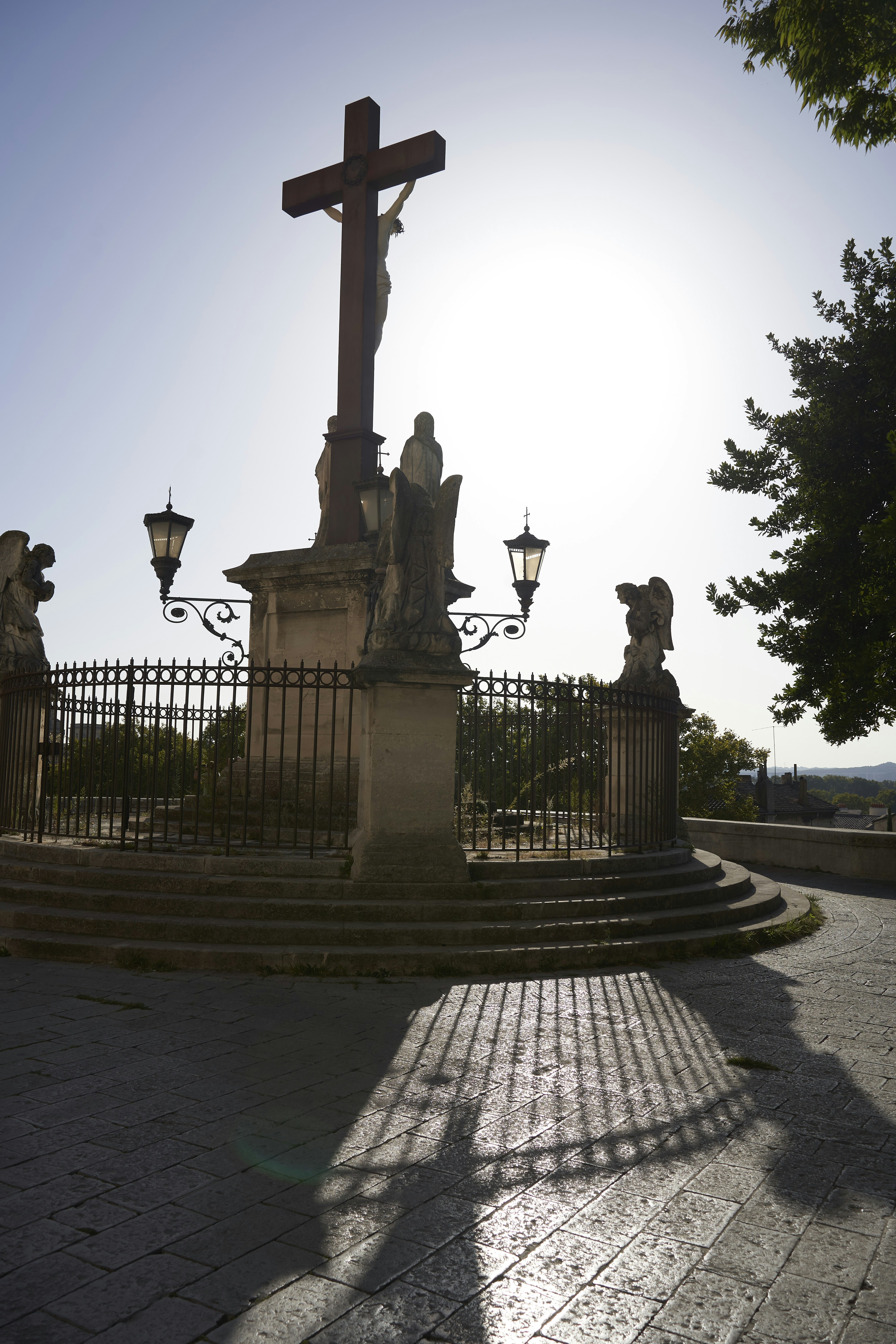 Stone cross and statues silhouetted against bright sky, casting intricate shadows on cobblestones in Avignon.