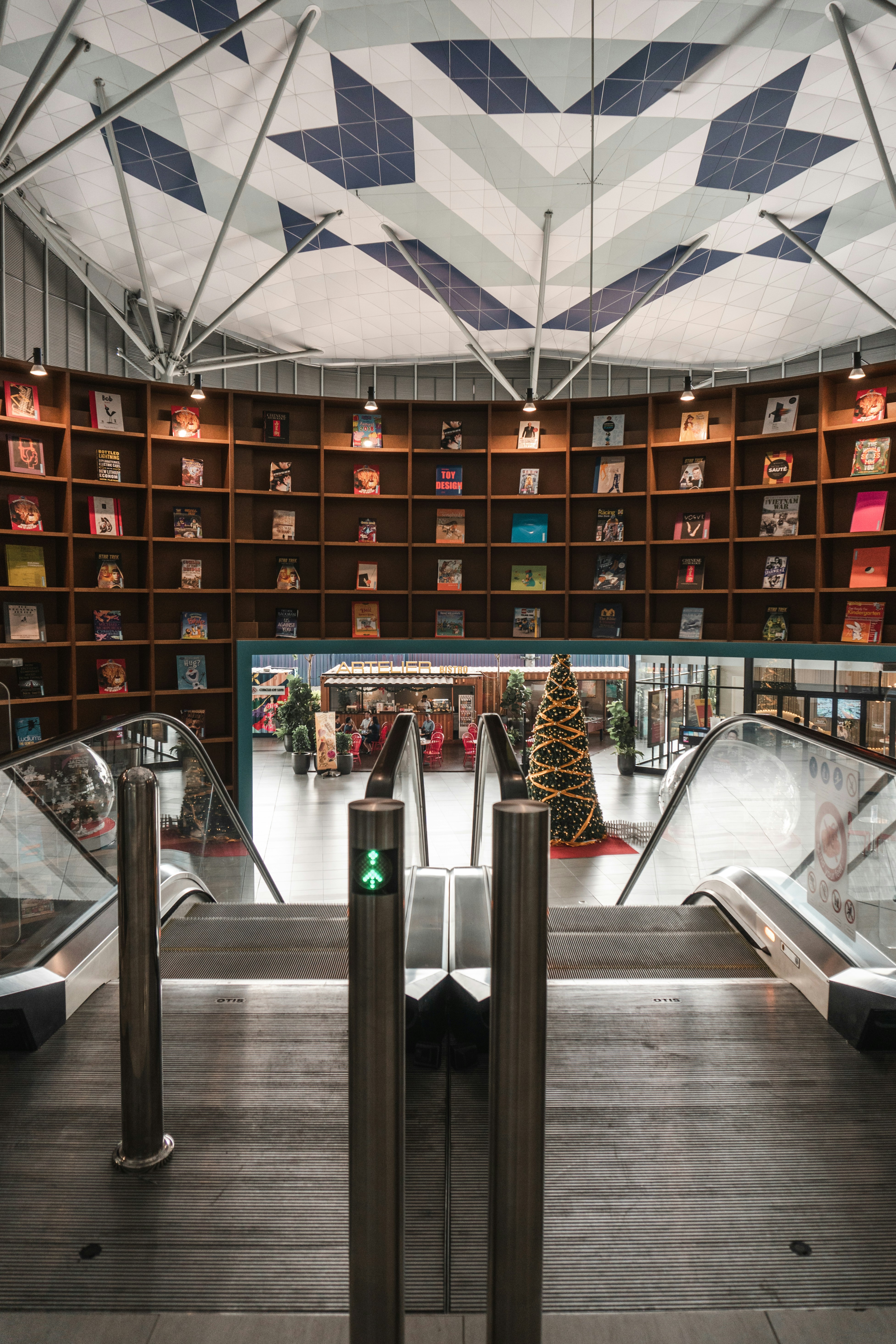 Escalator leading to a vibrant book display wall with a decorated Christmas tree below.