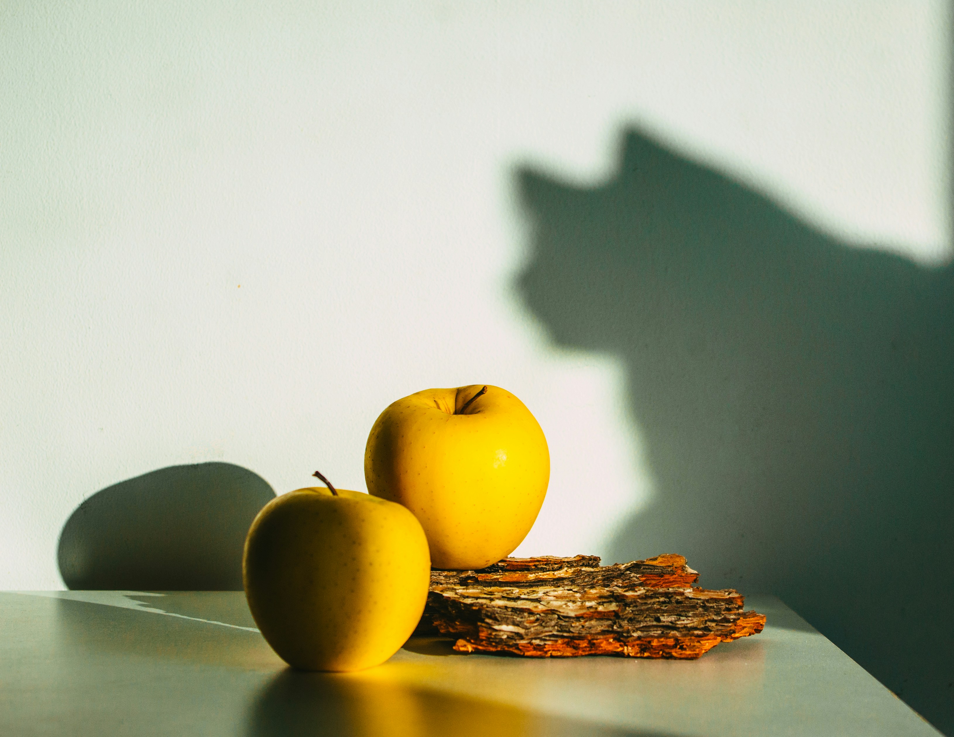 Two yellow apples sitting on top of a table next to a shadow of a cat ...