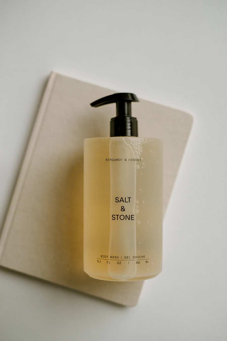 Translucent Salt & Stone body wash bottle with black pump resting on a beige book against a soft gray background. Minimal product styling with gentle shadows and warm tones.