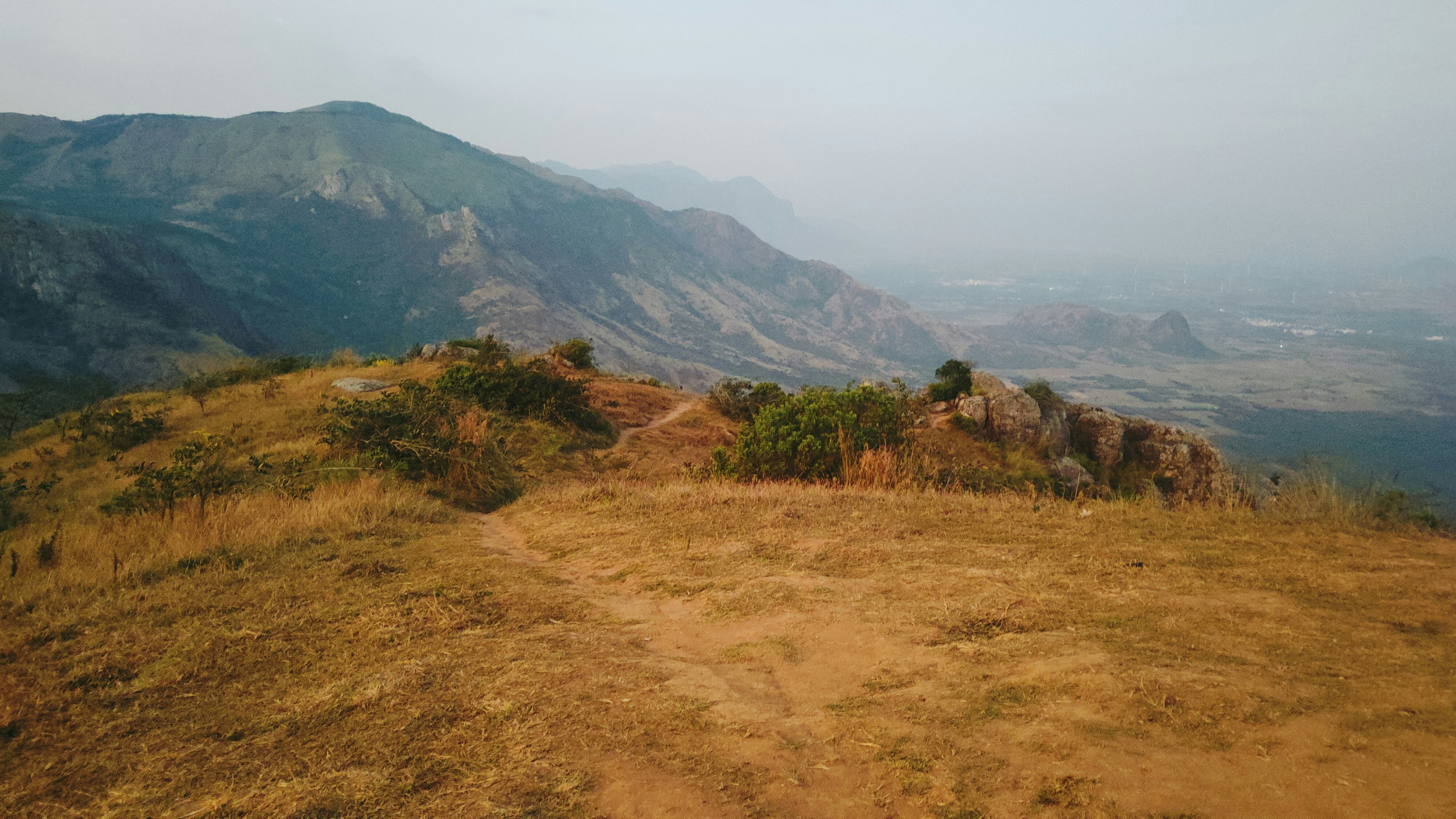 Dirt path winds across a sunlit hillside toward the hazy mountains on the distant horizon. Sparse shrubs and rocky outcrops punctuate the slope in this sunlit landscape photograph.