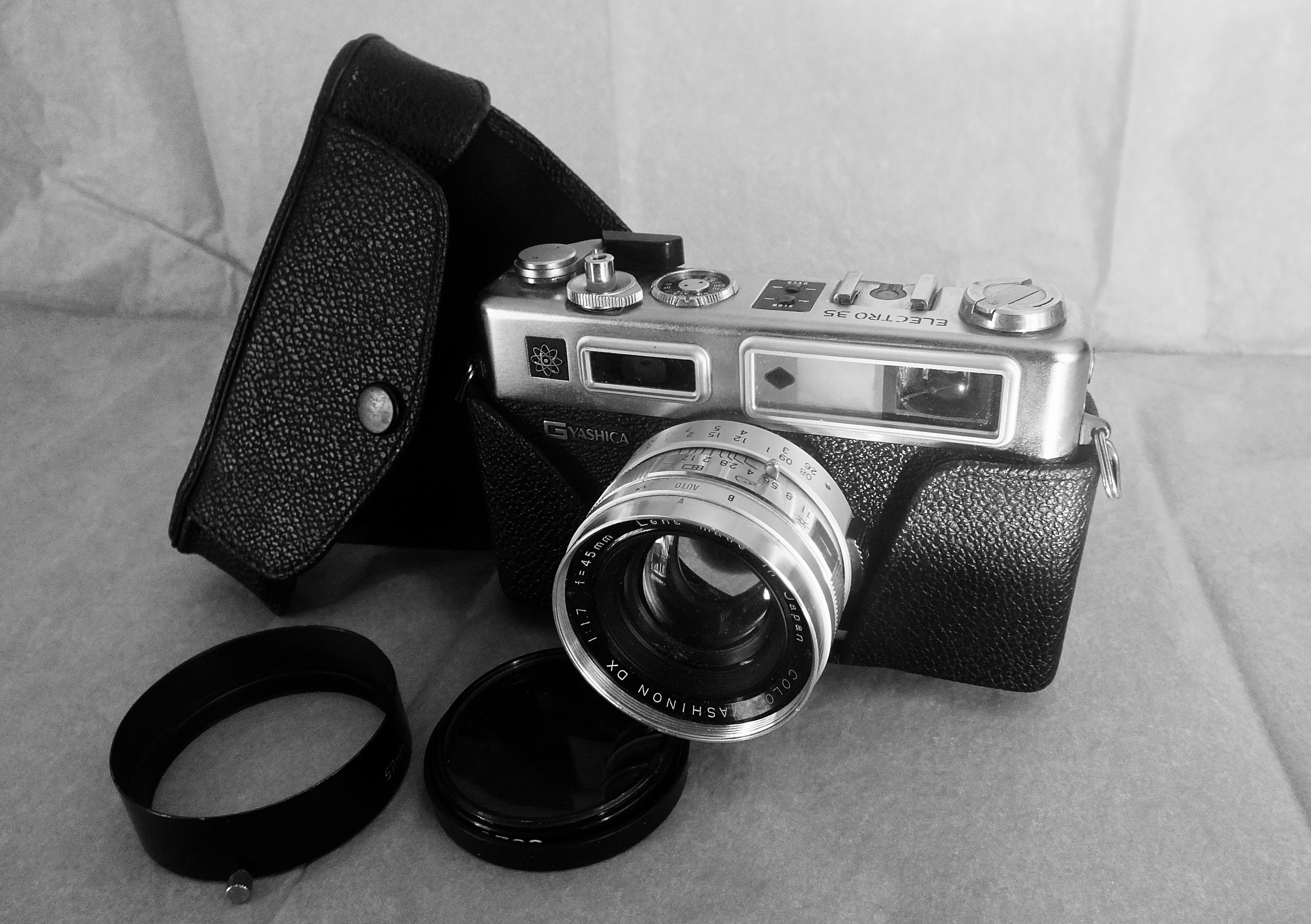 Monochrome photograph of a vintage rangefinder camera with a metal lens mounted and a black lens cap nearby. A leather strap and an extra ring lie beside it on a soft backdrop.