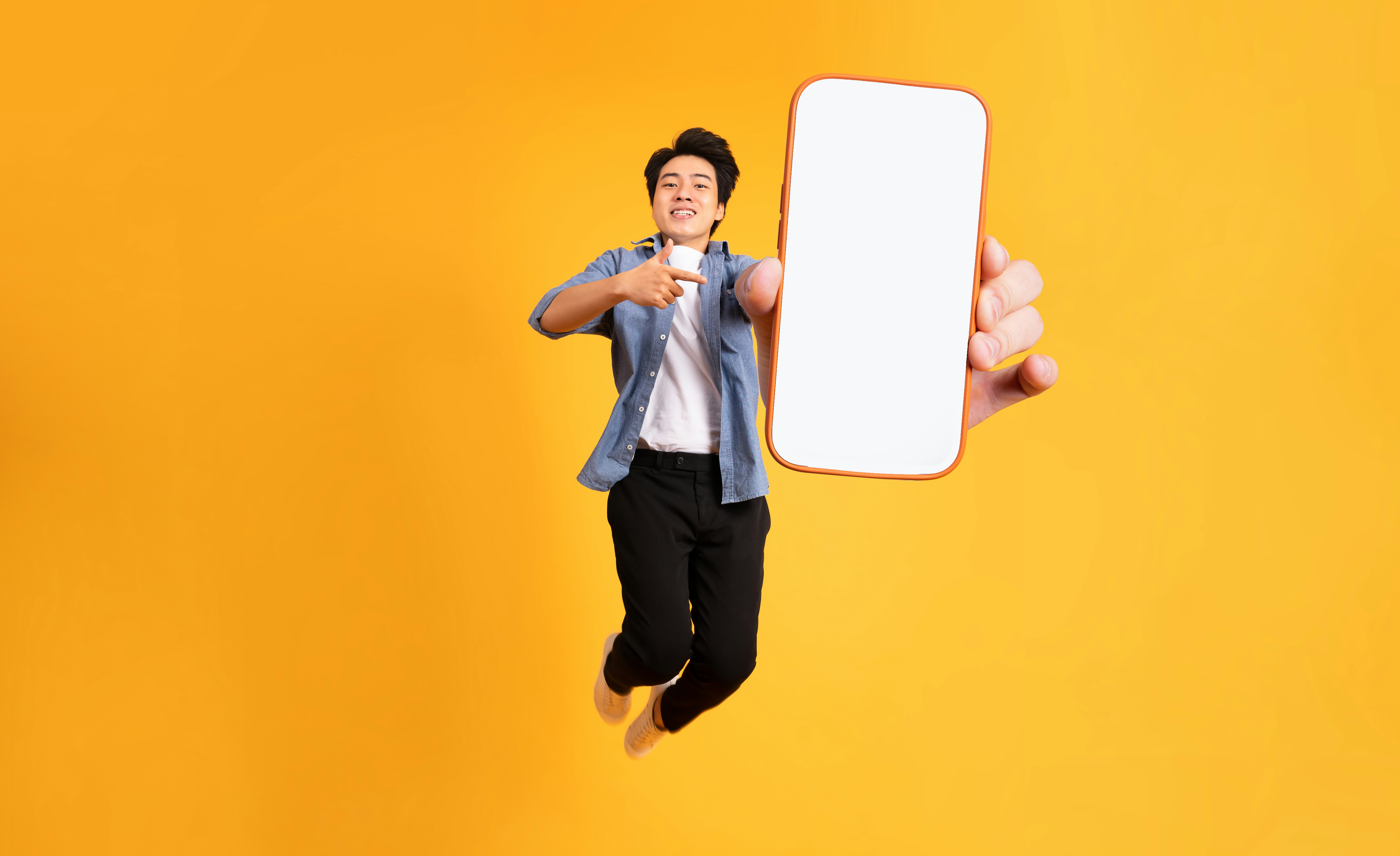 Young man joyfully leaping against a vibrant yellow backdrop, showcasing a blank smartphone screen for promotional purposes.
