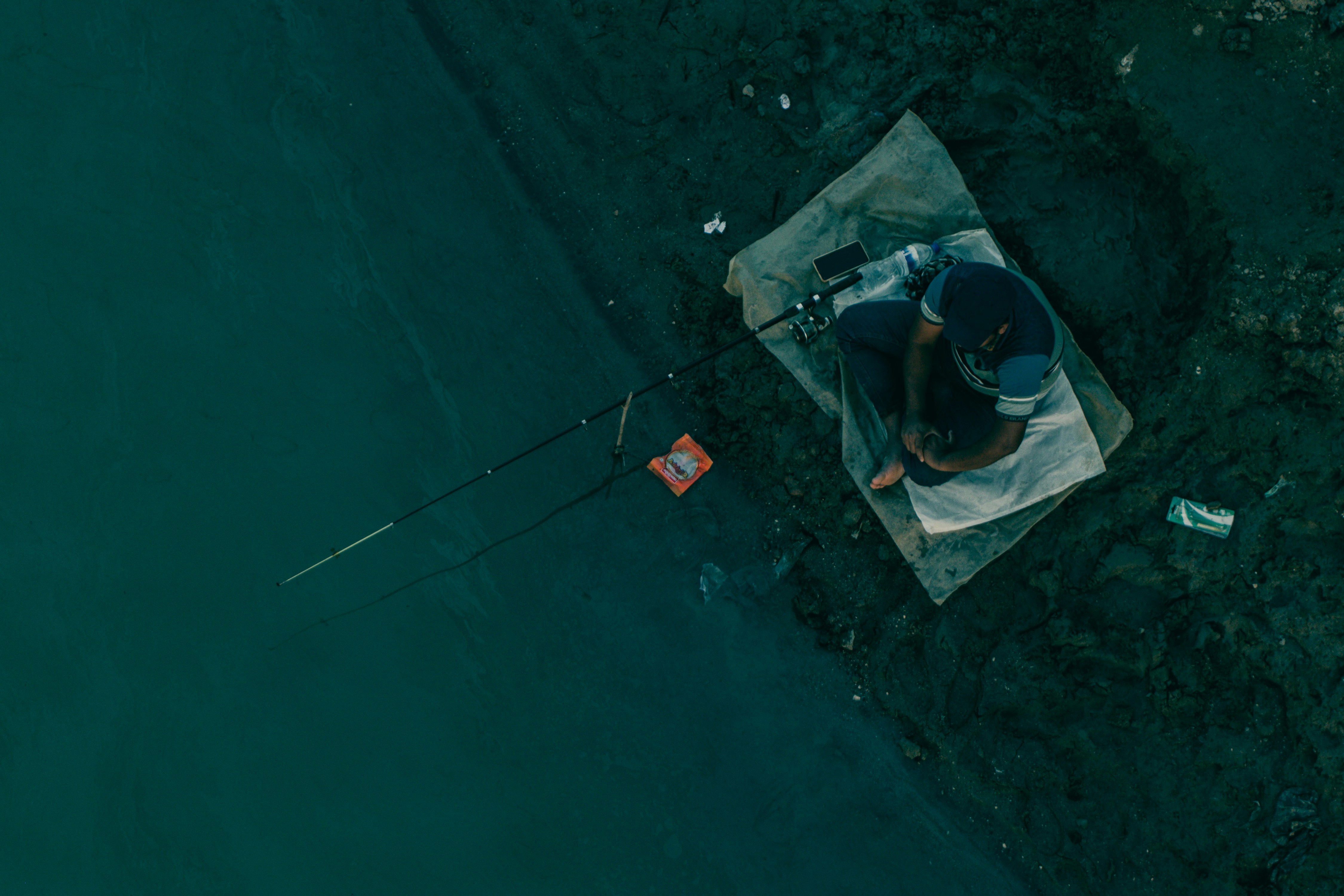 Person sitting on a riverbank with a fishing pole, viewed from above on a small platform.