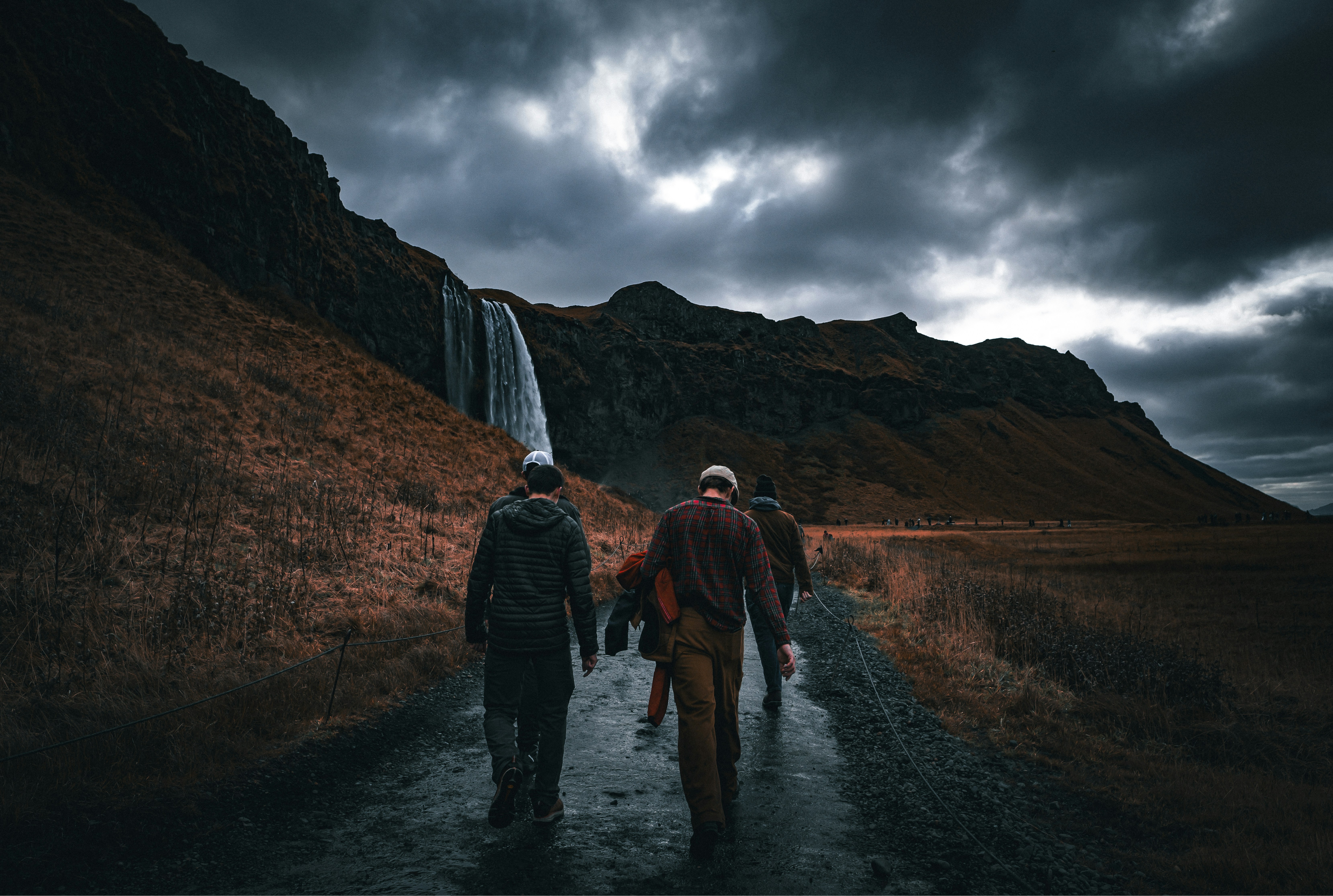 Hikers on a moody Icelandic trail toward a veiled waterfall under a stormy sky
