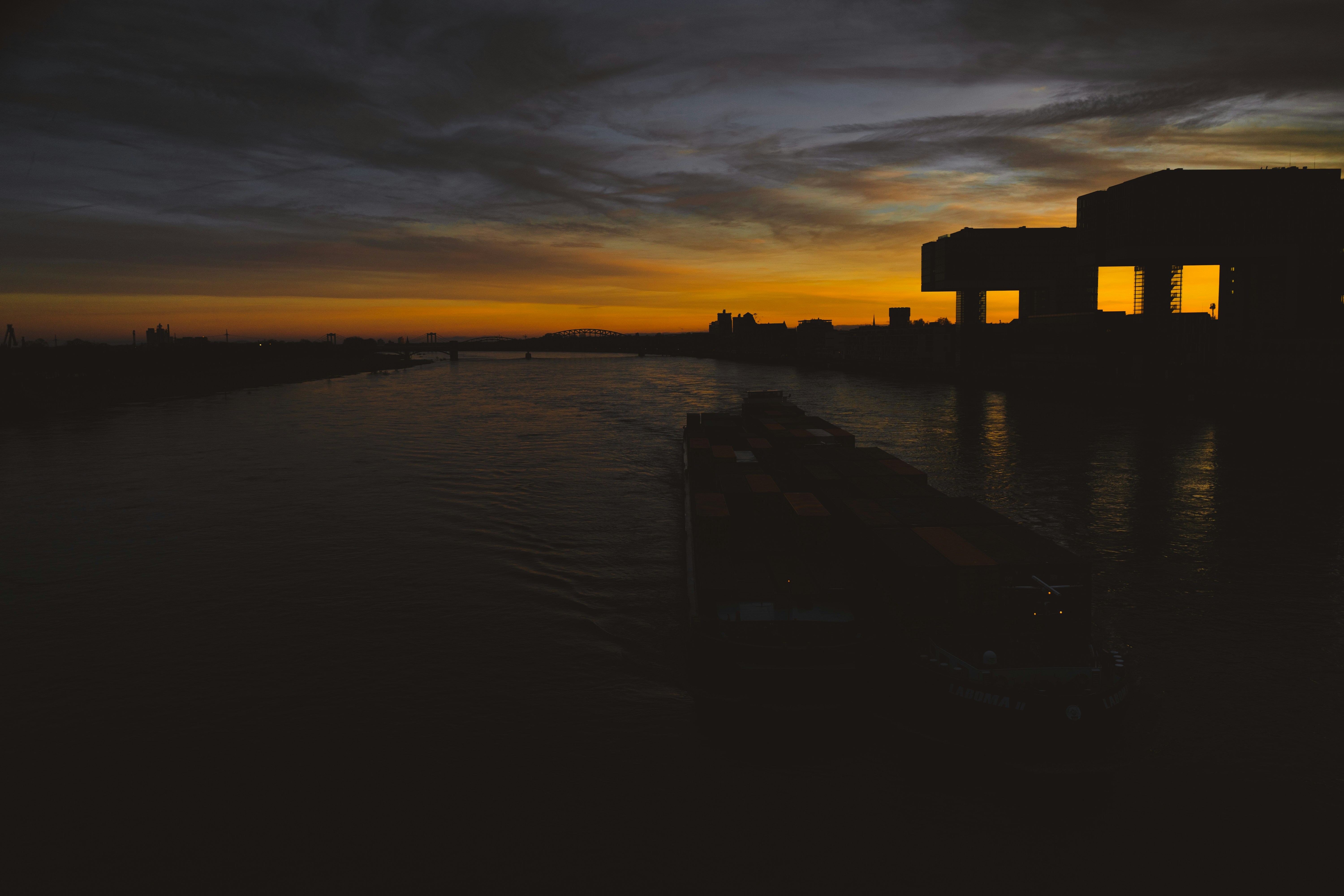 Silhouetted structures beside a calm river under a vibrant sunset sky.