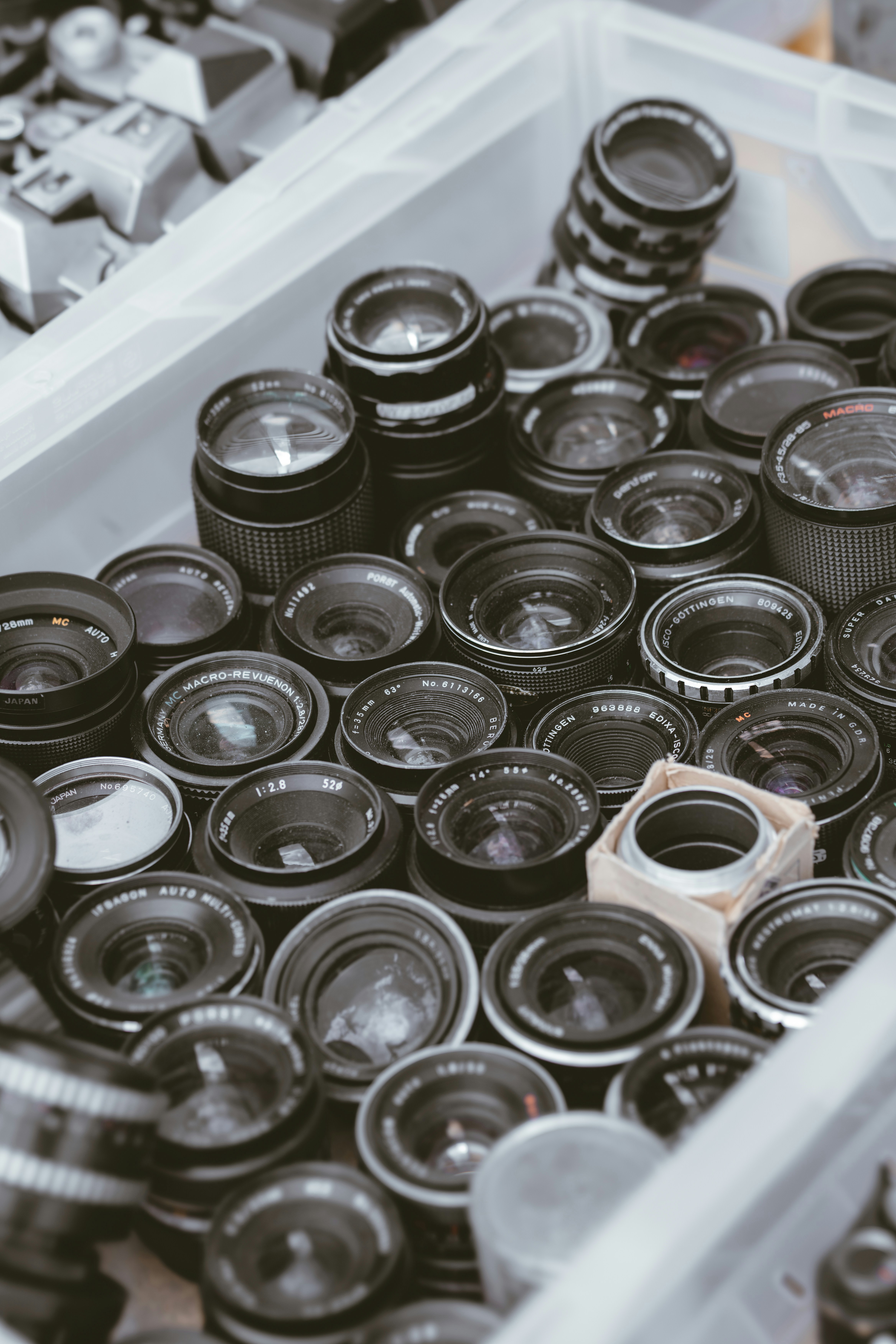 An assortment of vintage camera lenses displayed in a clear container, showcasing various shapes and sizes. The collection highlights the diversity of photographic equipment.
