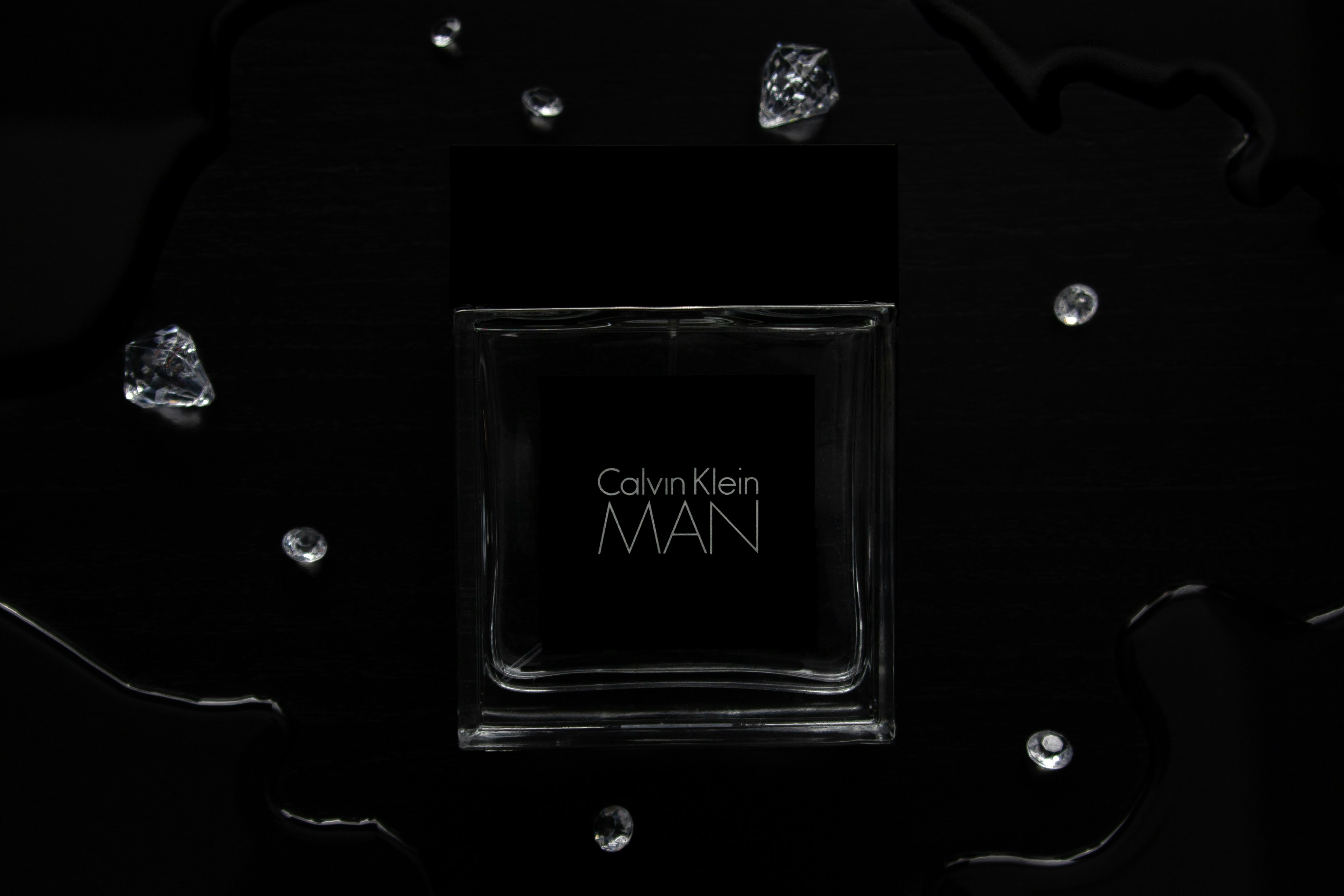 Calvin Klein MAN fragrance bottle surrounded by scattered gemstones on a dark surface, emphasizing luxury and sophistication.