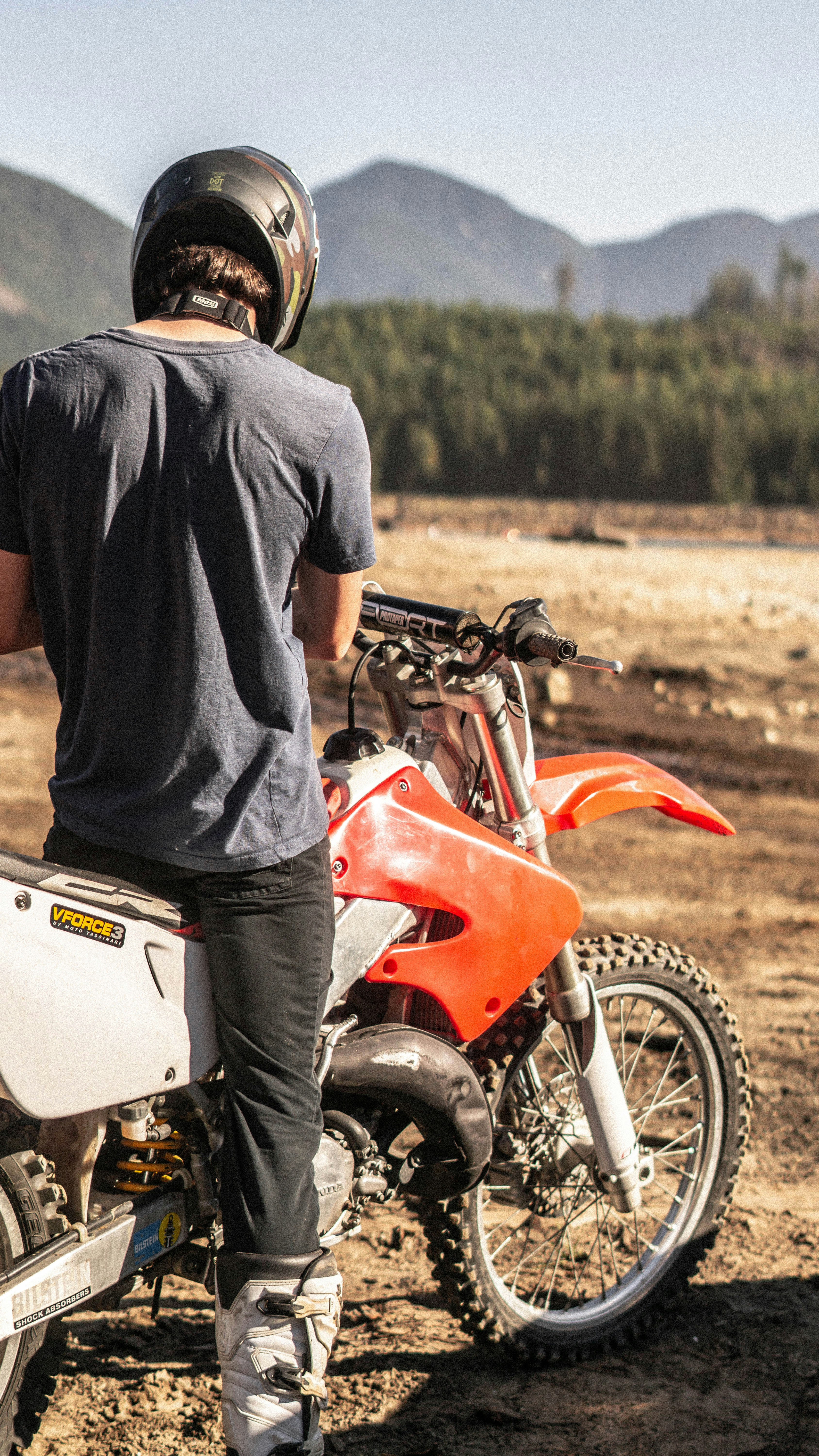 A rider in a helmet stands beside a dirt bike, preparing for an adventure in a rugged terrain under a clear sky.