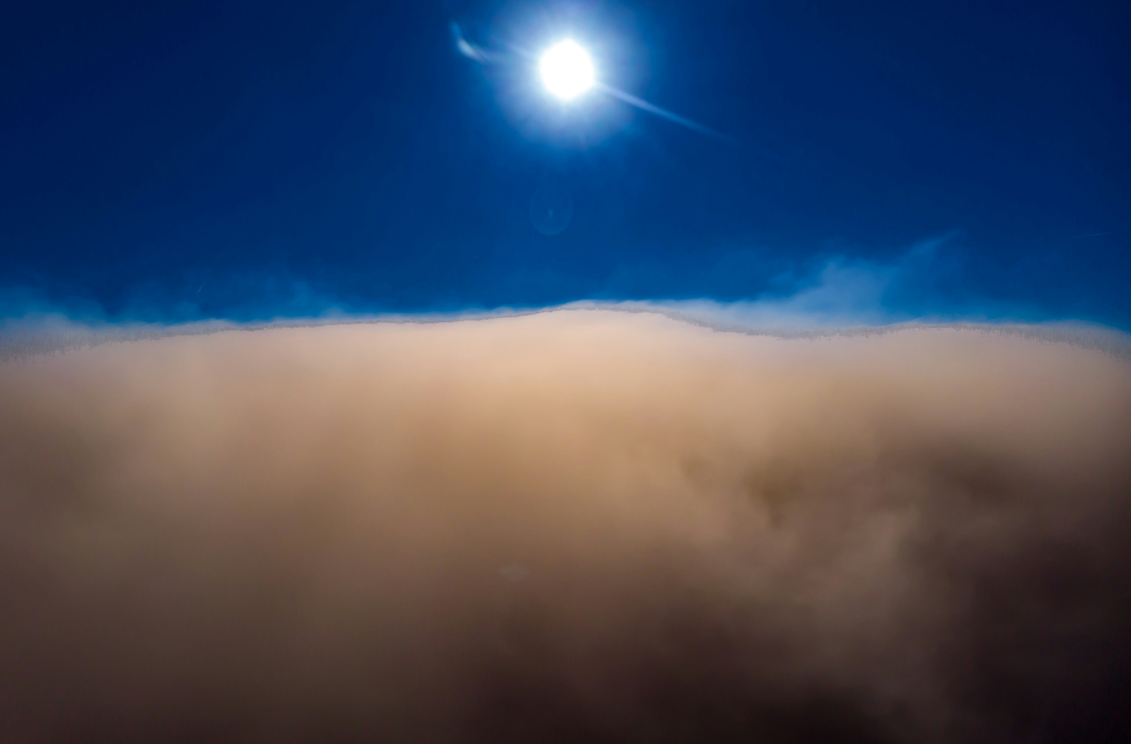 A serene aerial view of a thick, fluffy cloud layer under a clear blue sky with the sun glaring brightly above. The contrast between the warm tones of the clouds and the cool blue sky creates a dramatic and tranquil atmosphere