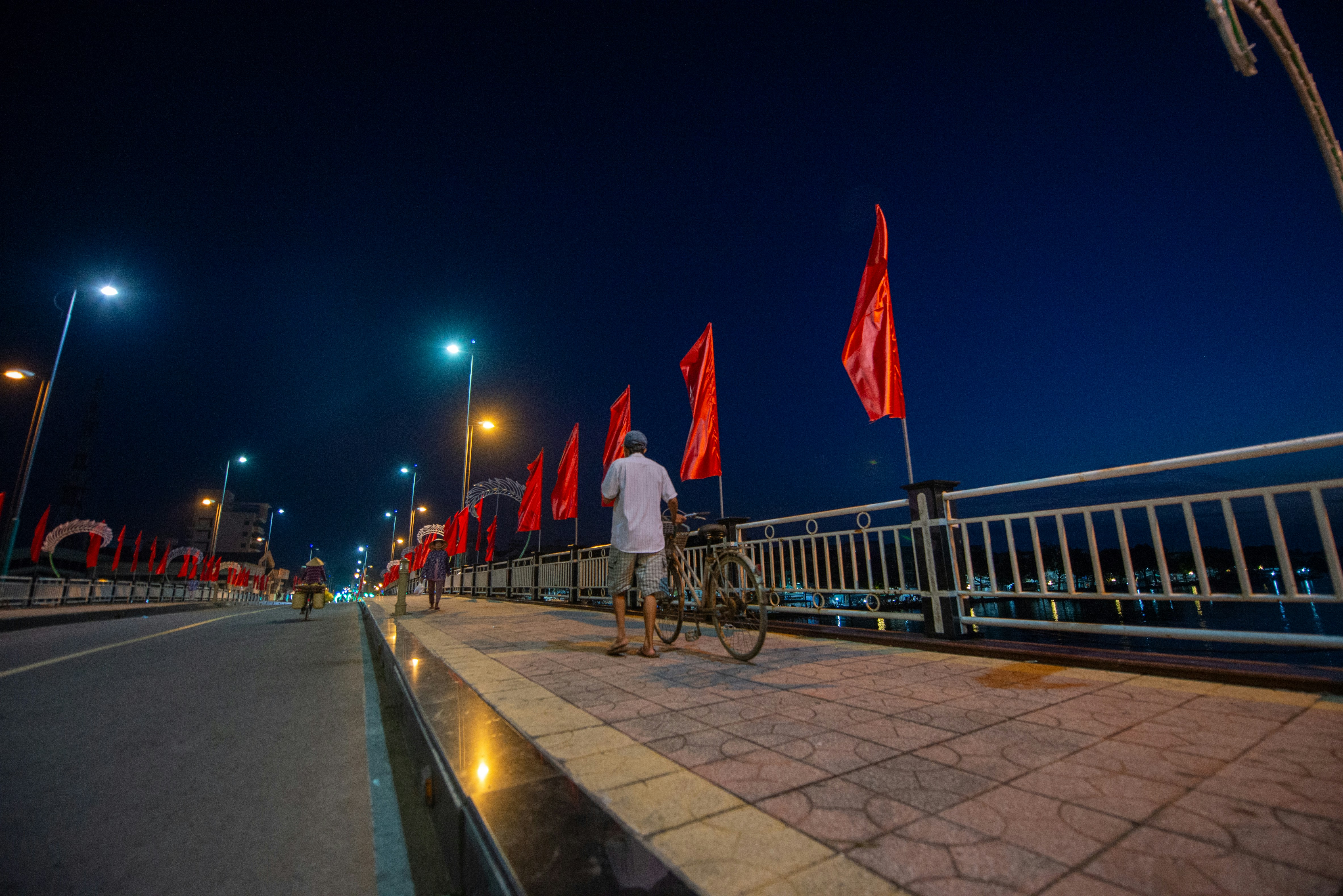 Midnight Pedestrian with Bicycle Under Crimson Flags on a Night Pier