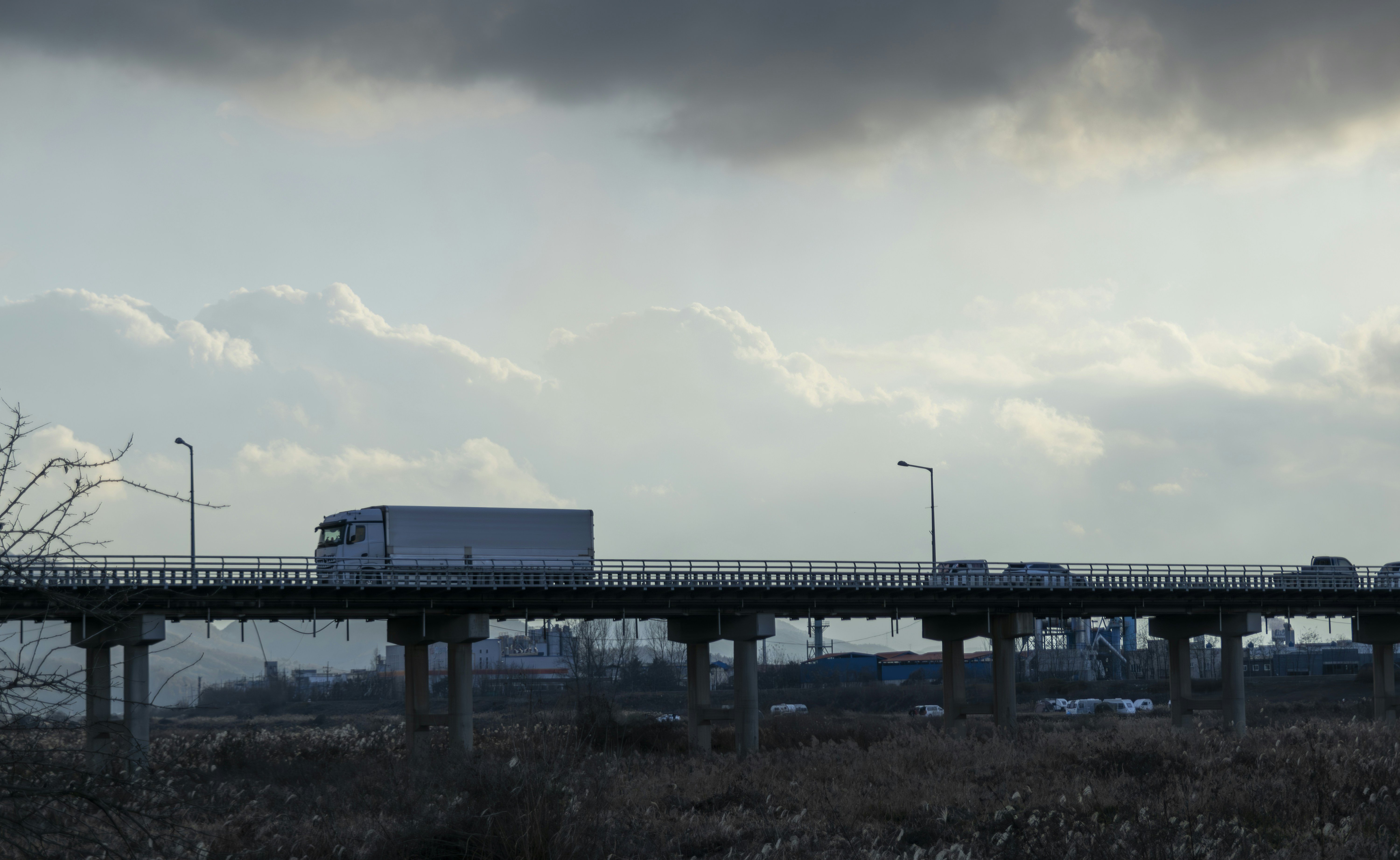 A transport truck traverses a bridge, silhouetted against a moody sky with scattered clouds. The scene captures the intersection of infrastructure and nature.