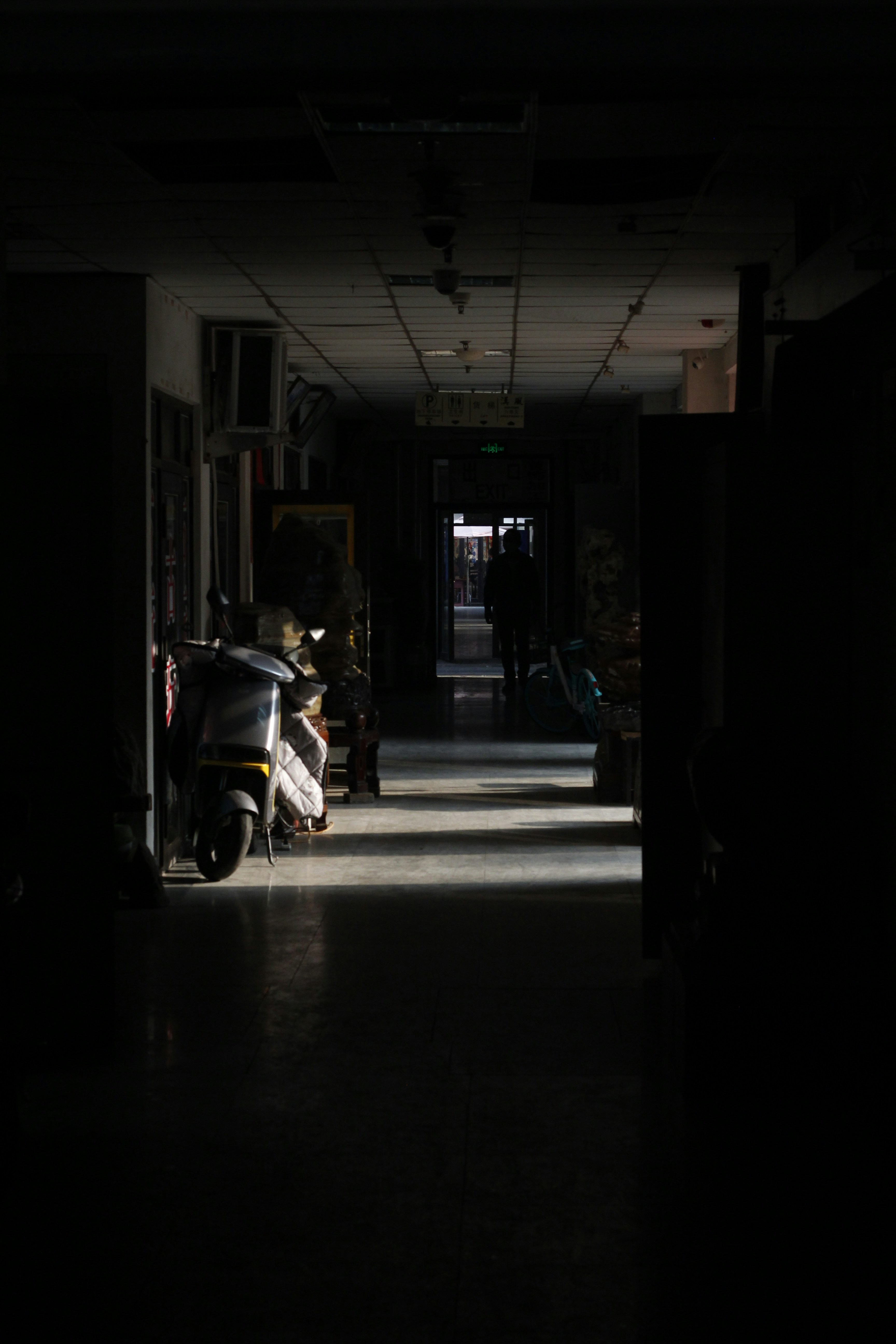 An underexposed and dark long indoor corridor with just few rays of light coming from the right side, lighting an electric scooter parked next to the left wall