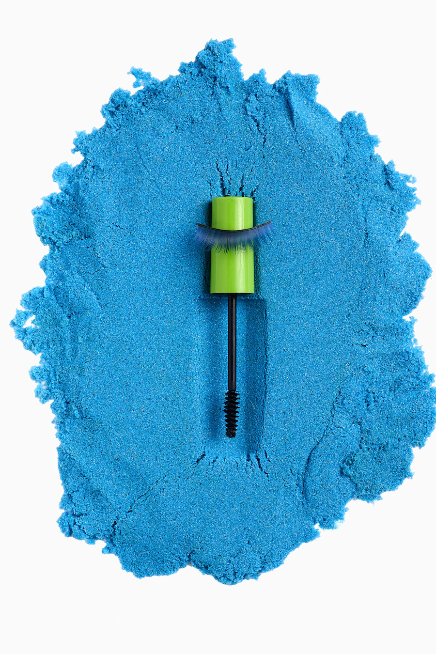 A lime green mascara wand stands upright in a bright blue powder splash on a white background.
