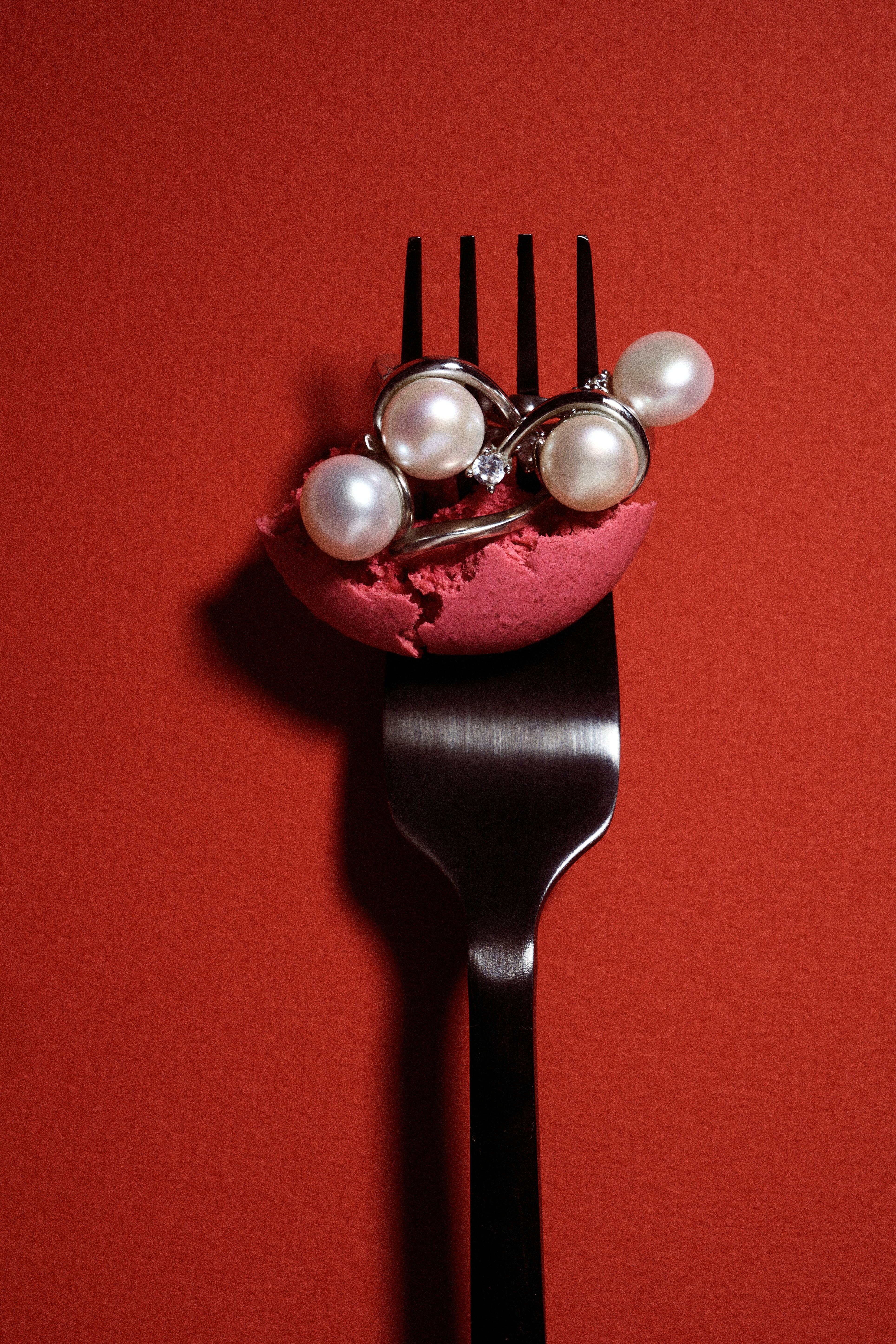 Photograph of a pink macaron topped with pearl jewelry balanced on a black fork against a red background.