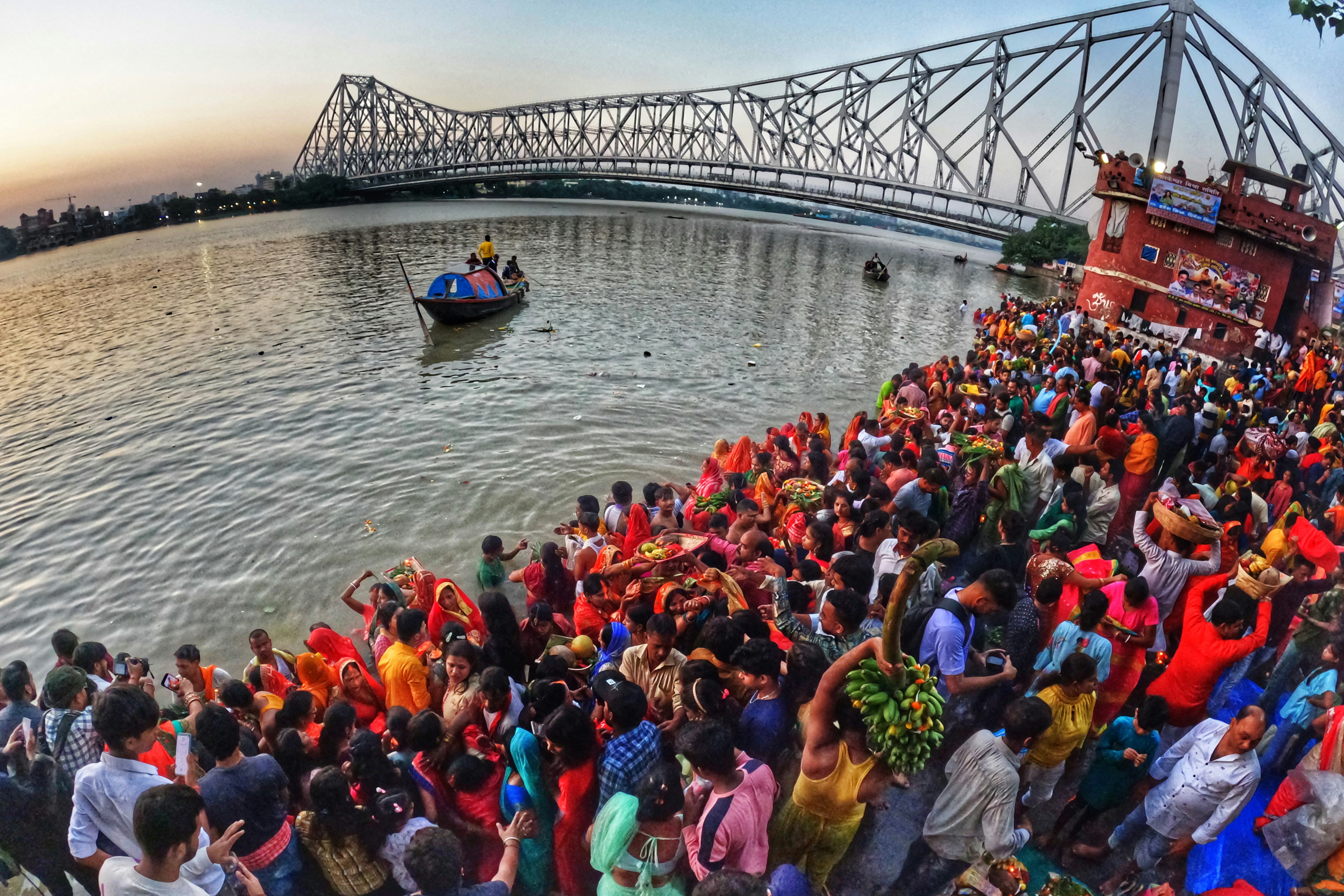 Vibrant crowd fills the riverfront beneath a steel truss bridge at dusk. A red stage anchors the right bank while a small boat glides across the calm water.