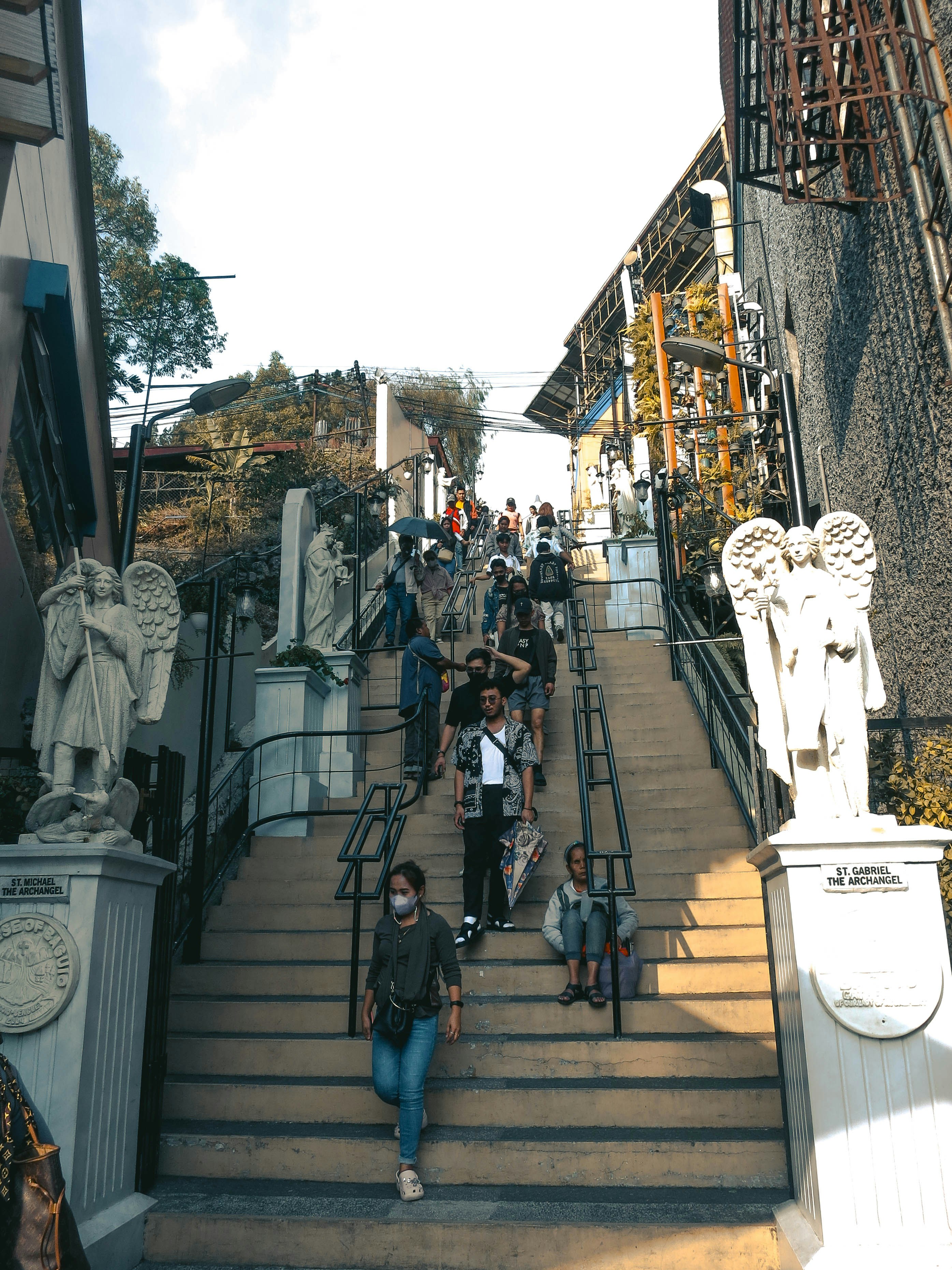 Crowd navigating a staircase flanked by angelic statues, leading to a vibrant cultural space. The scene captures the blend of art and daily life.