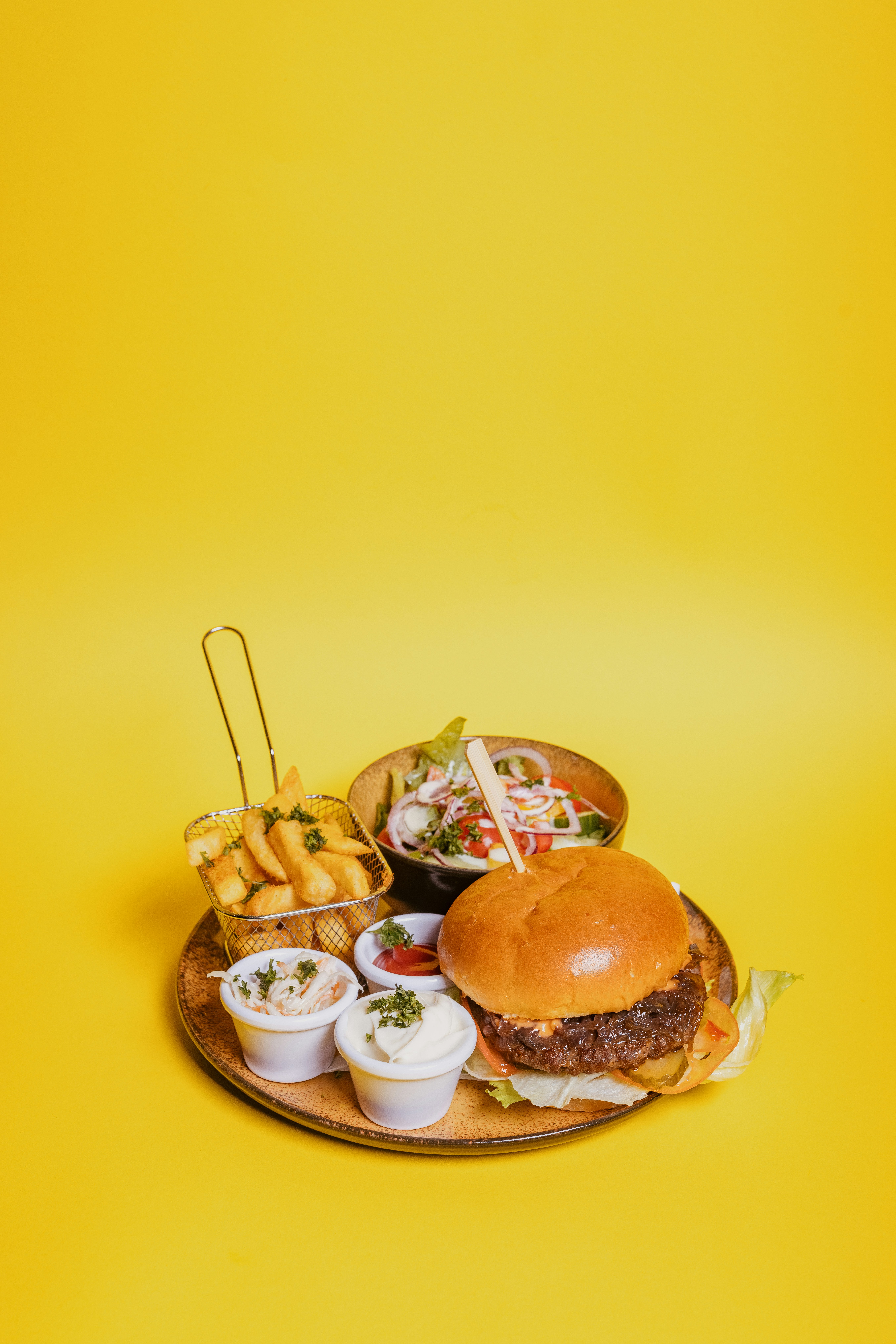 Burger platter with fries, salad, and sauces on a vibrant yellow background.