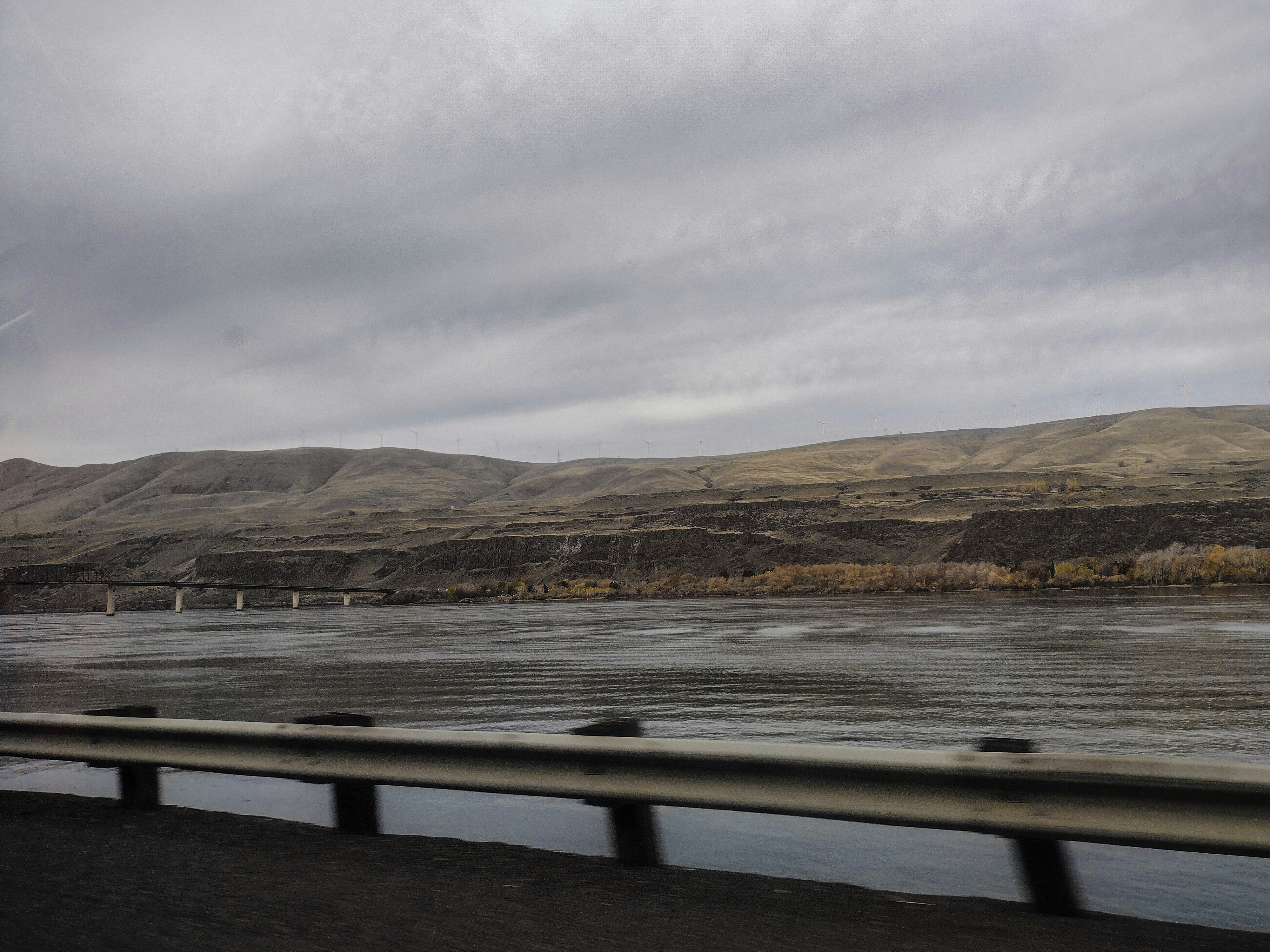 Overcast riverscape with a guardrail in the foreground and distant rolling hills across calm water.