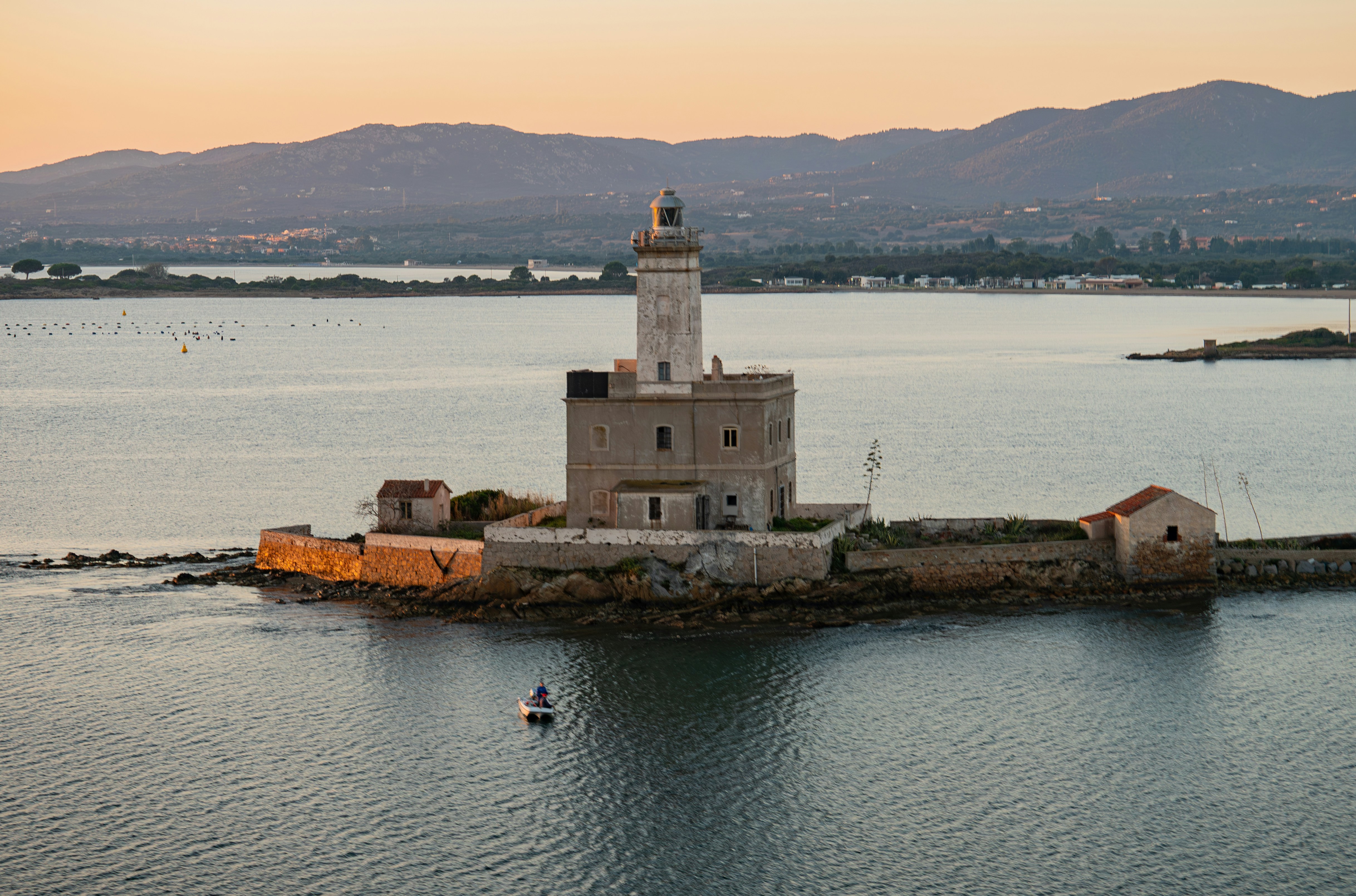 Lighthouse on a small island surrounded by calm waters at sunset, with distant mountains under a pastel sky.