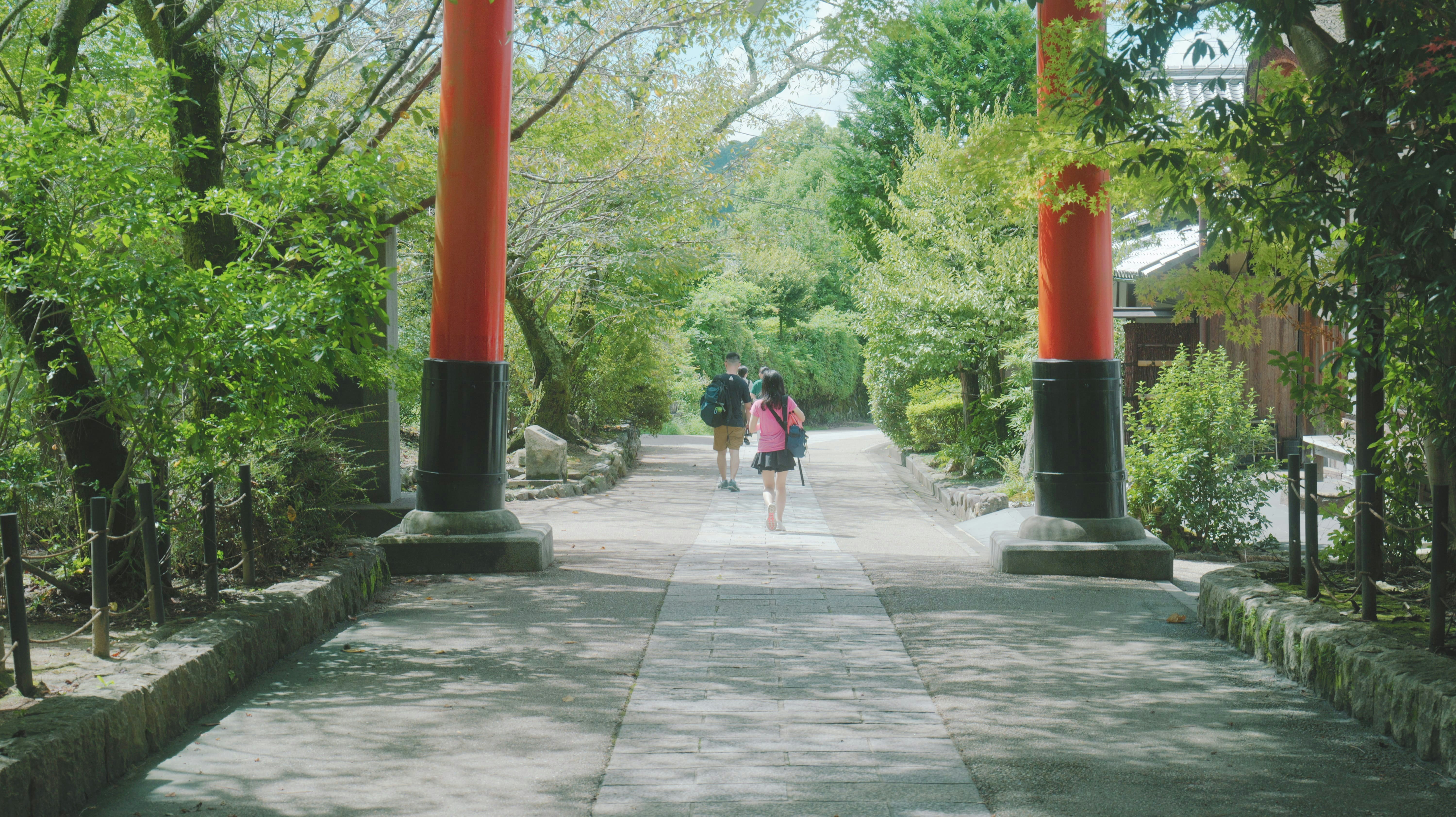 Two individuals strolling along a serene pathway framed by lush greenery and traditional red torii gates. The scene captures a tranquil moment in a cultural landscape.
