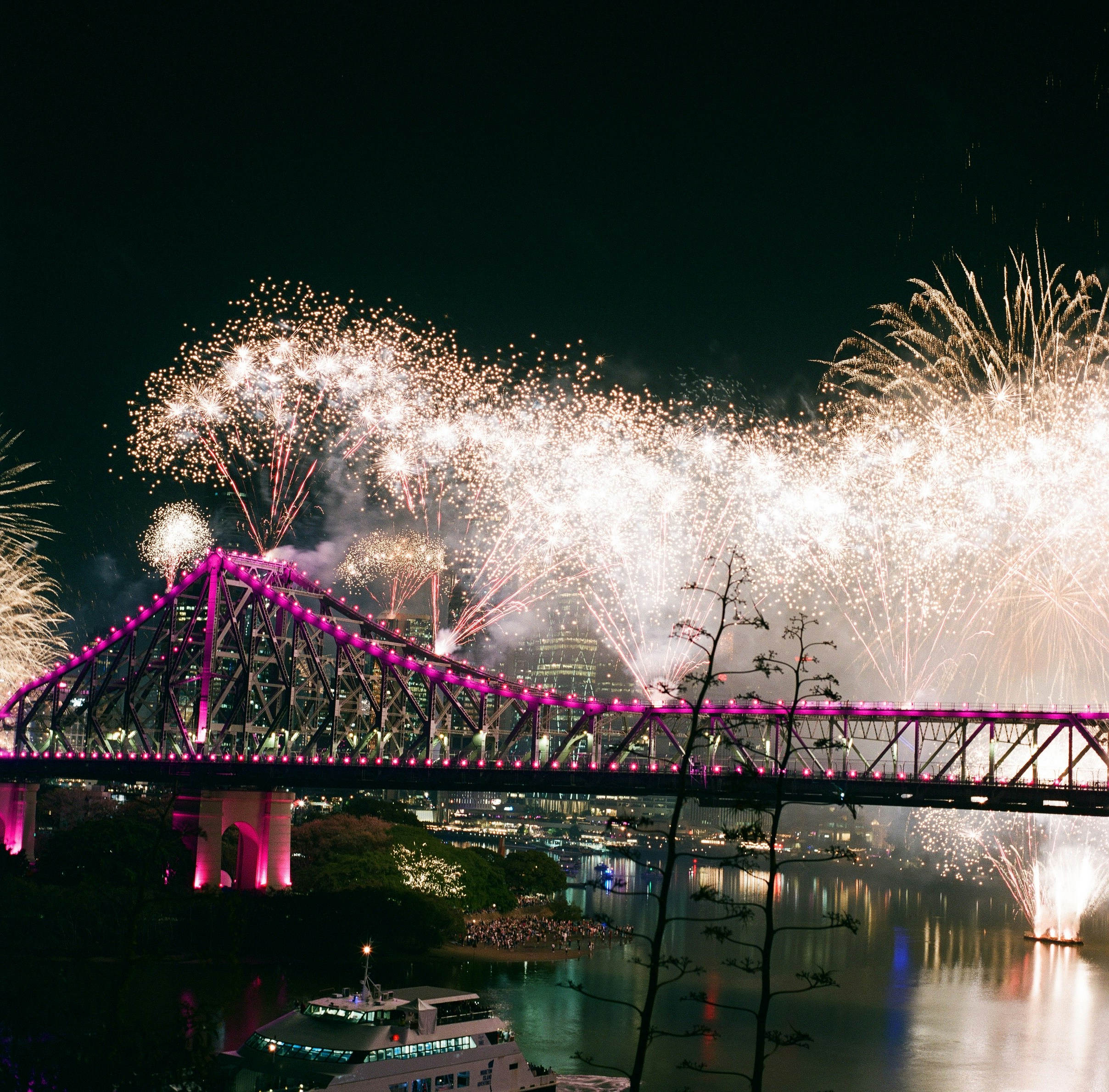 Night photograph of a purple-lit bridge spanning a calm river as fireworks explode above the city skyline.