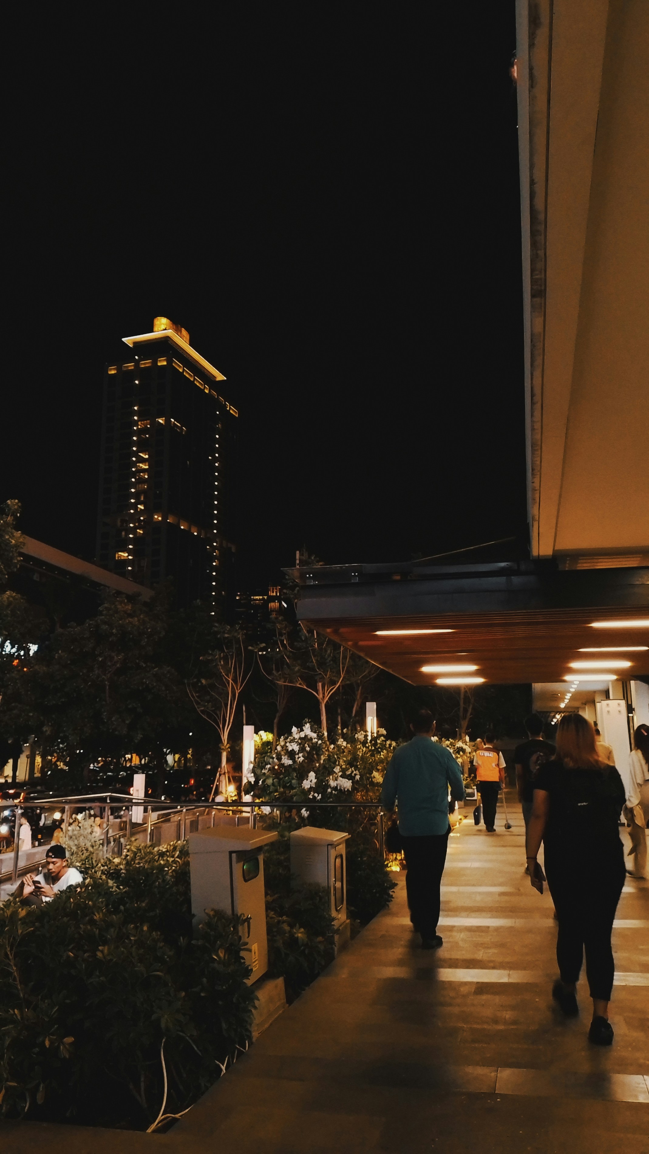 Nighttime urban walkway bathed in warm lighting, with a tall illuminated tower to the left and pedestrians along a planted railing.