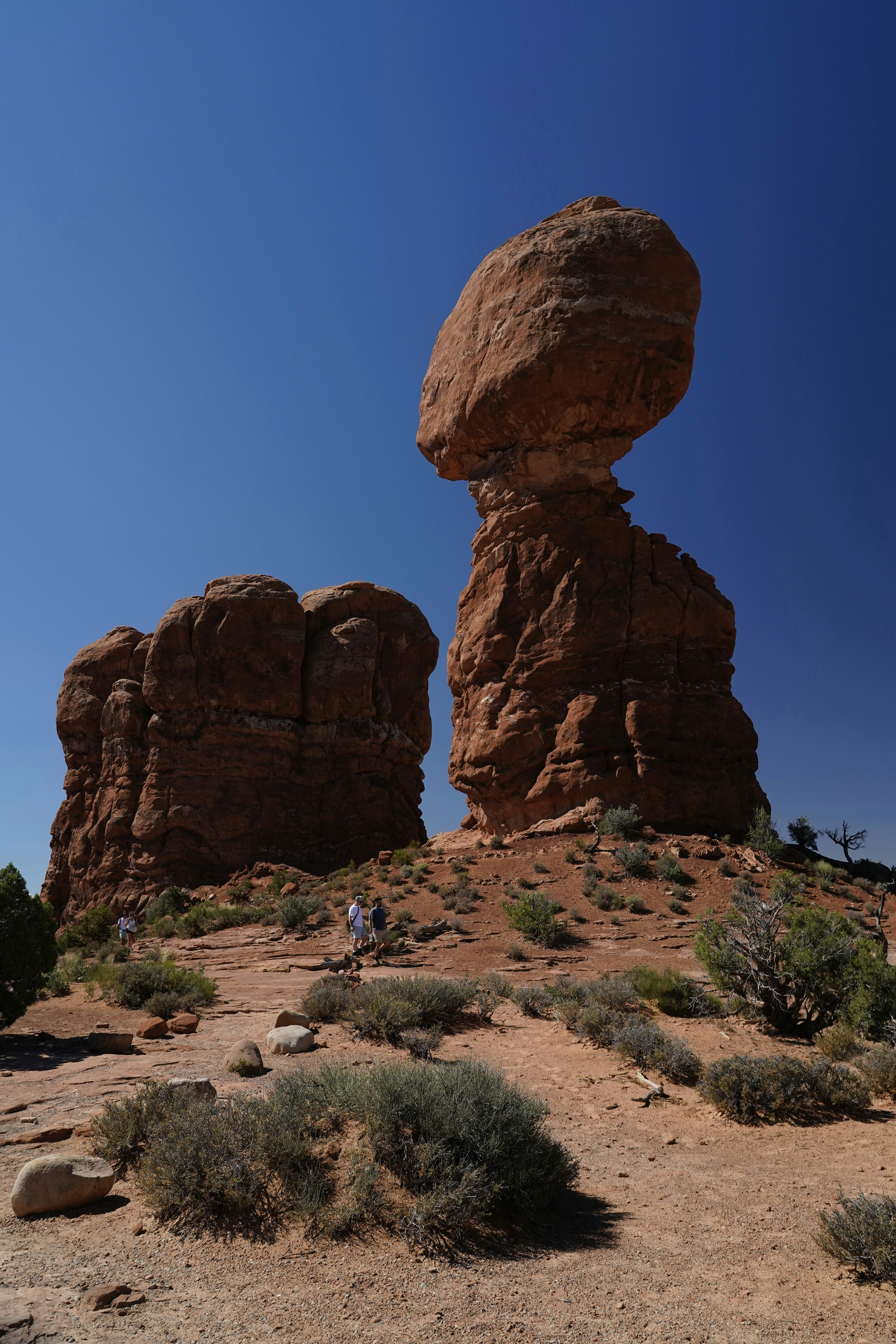 A towering rock formation, with a large boulder precariously balanced atop a slender pillar, set against a clear blue sky. Visitors explore the rugged terrain below.