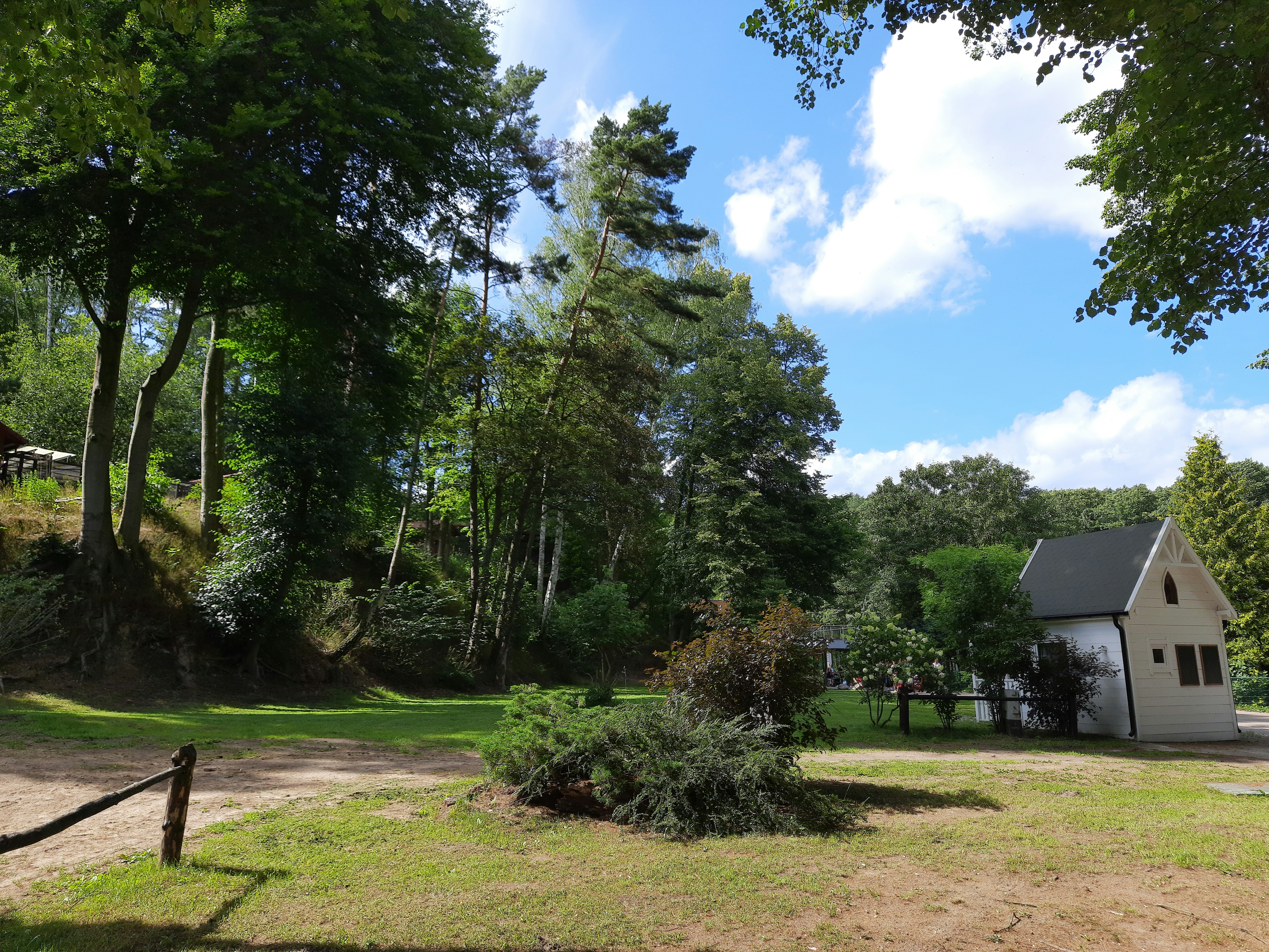 Sunlit park scene with a small white cottage to the right, framed by tall trees and a grassy clearing. A dirt path winds through the left foreground.