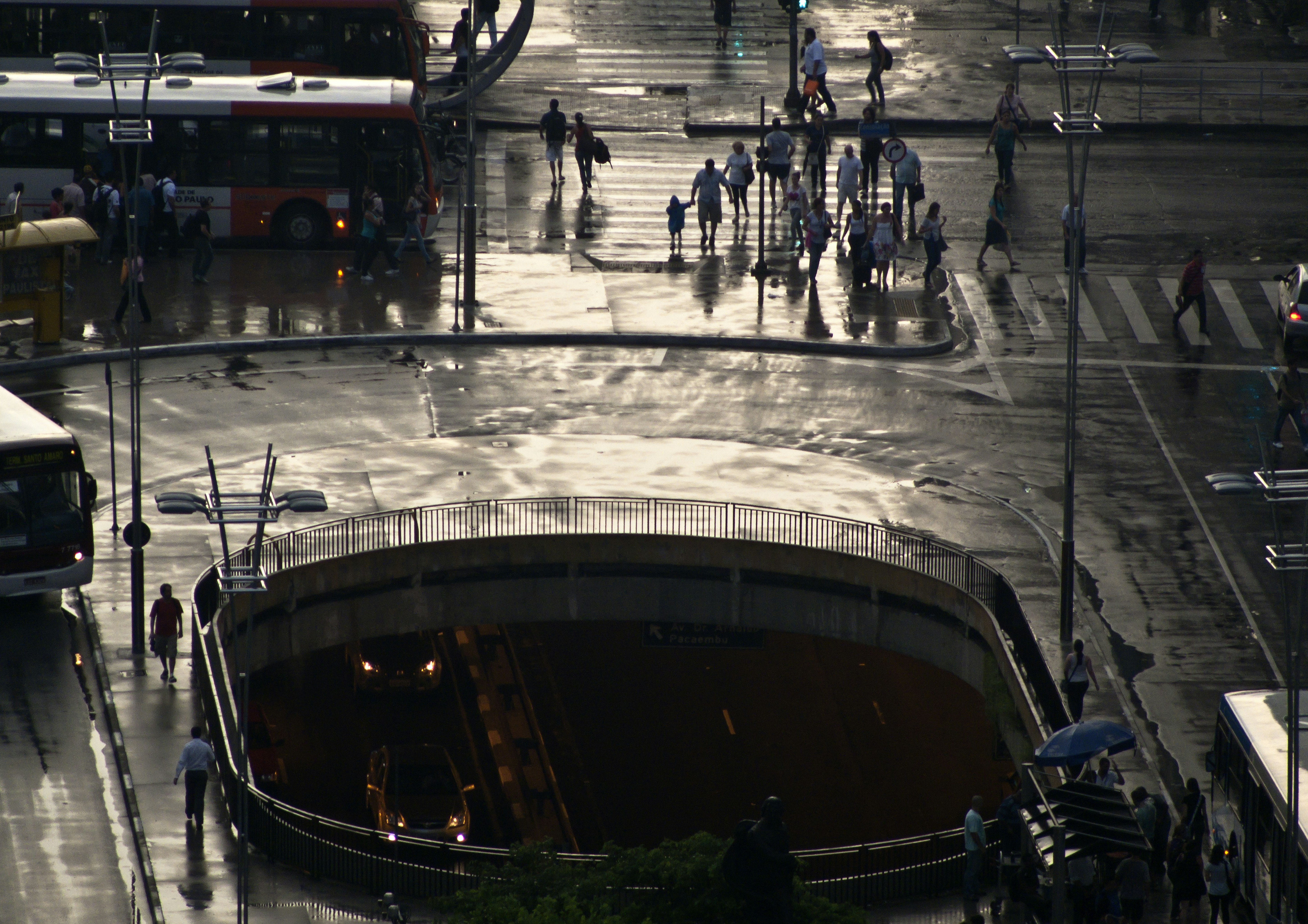 A rain-soaked city square photographed from above, centered on a circular underground entrance framed by wet reflections and scattered pedestrians.