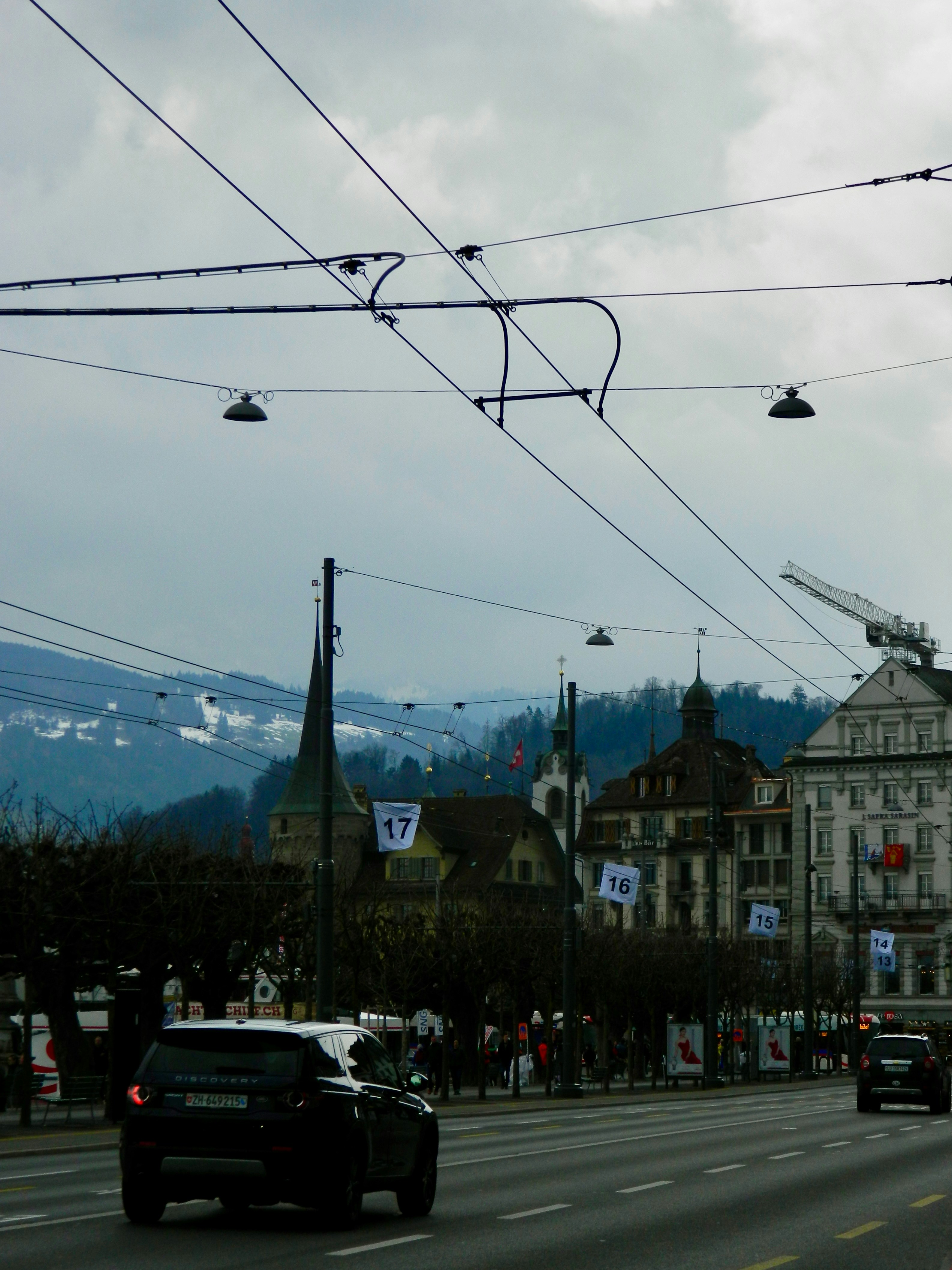 Urban street scene with overhead tram cables crisscrossing the sky, passing cars, and distant hills under a cloudy sky.