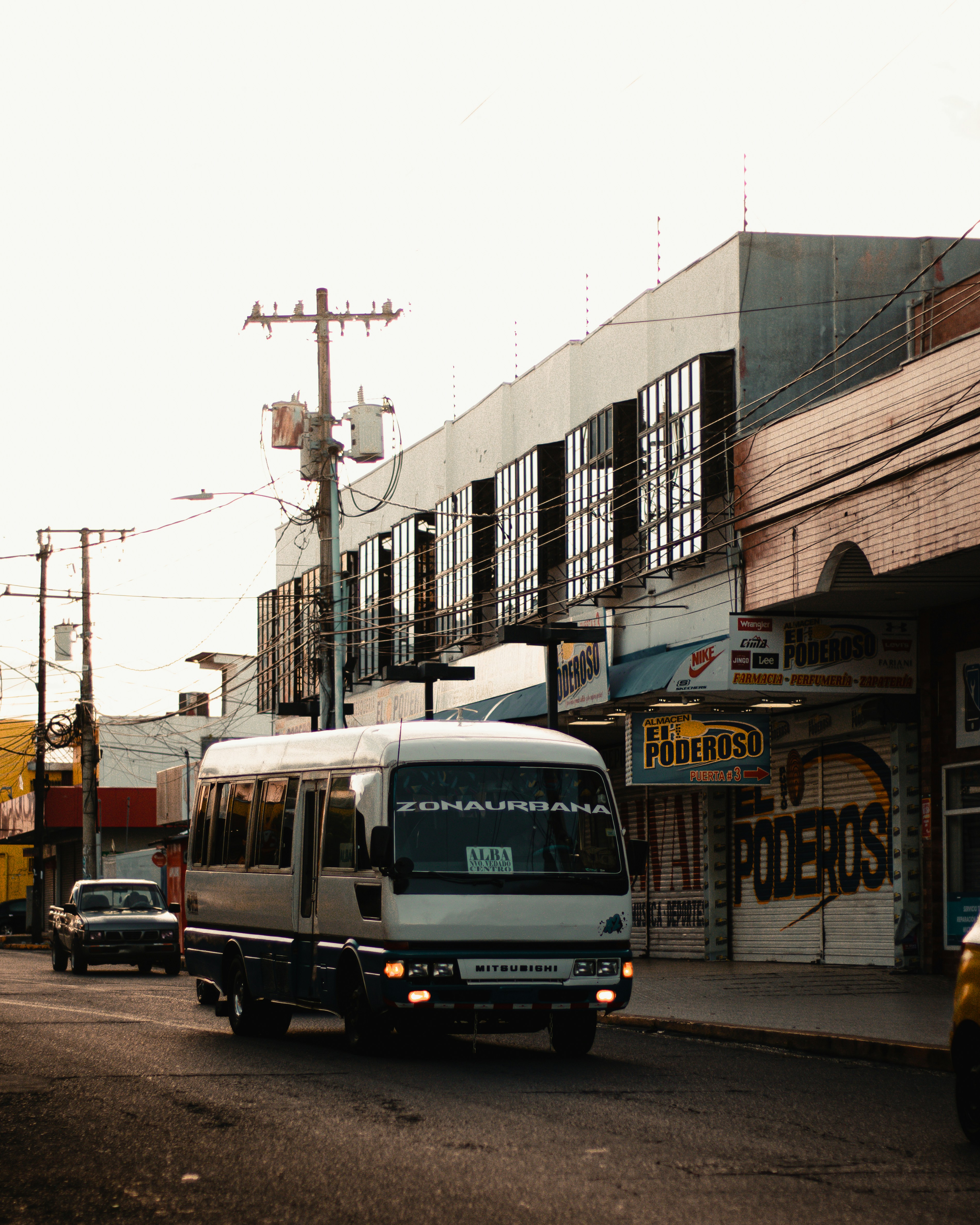 White Minibus Navigates a Sunlit Avenida Flanked by Shuttered Shops