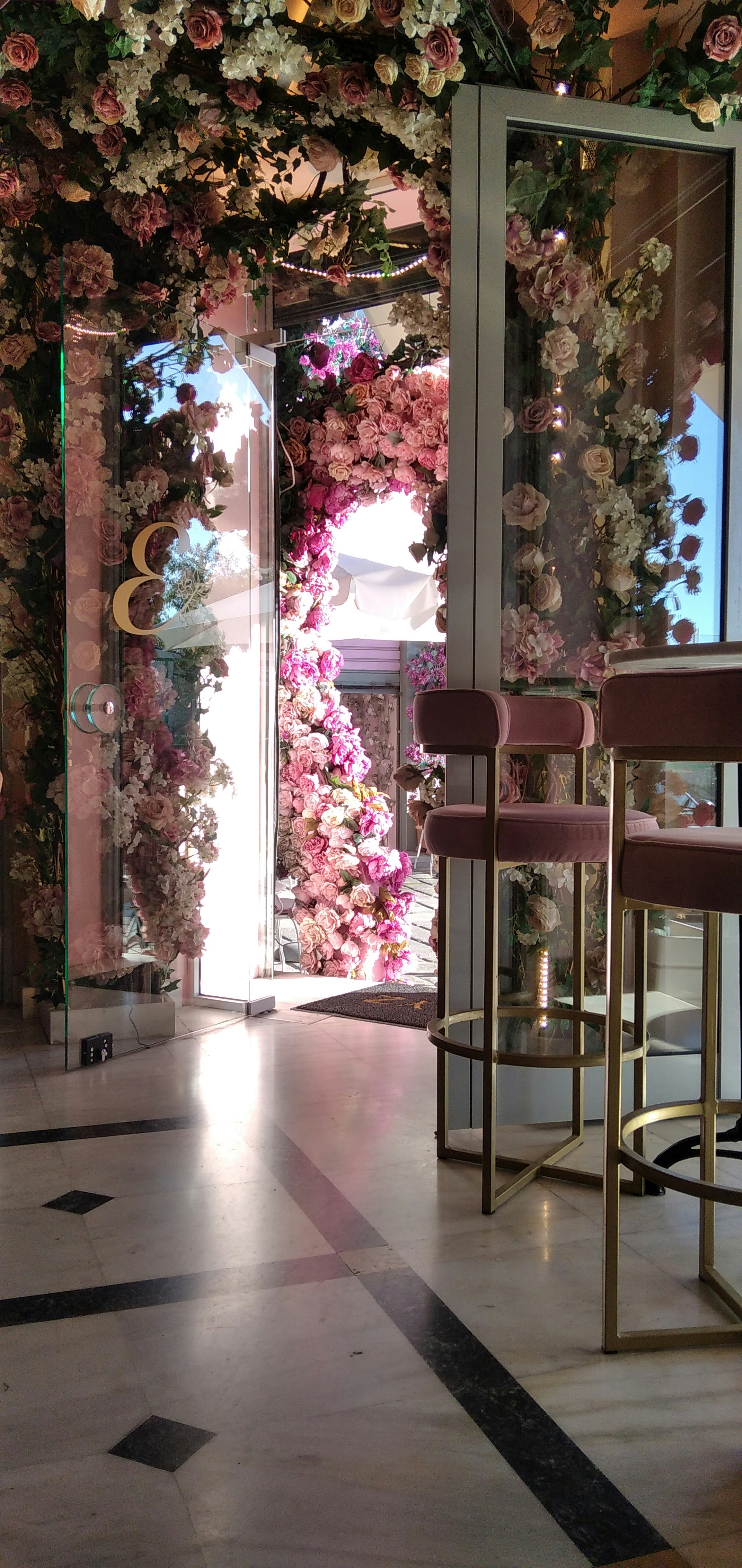 A photograph of a rose-filled arch framing a glass door in a chic lounge, with pink velvet stools and gold accents.