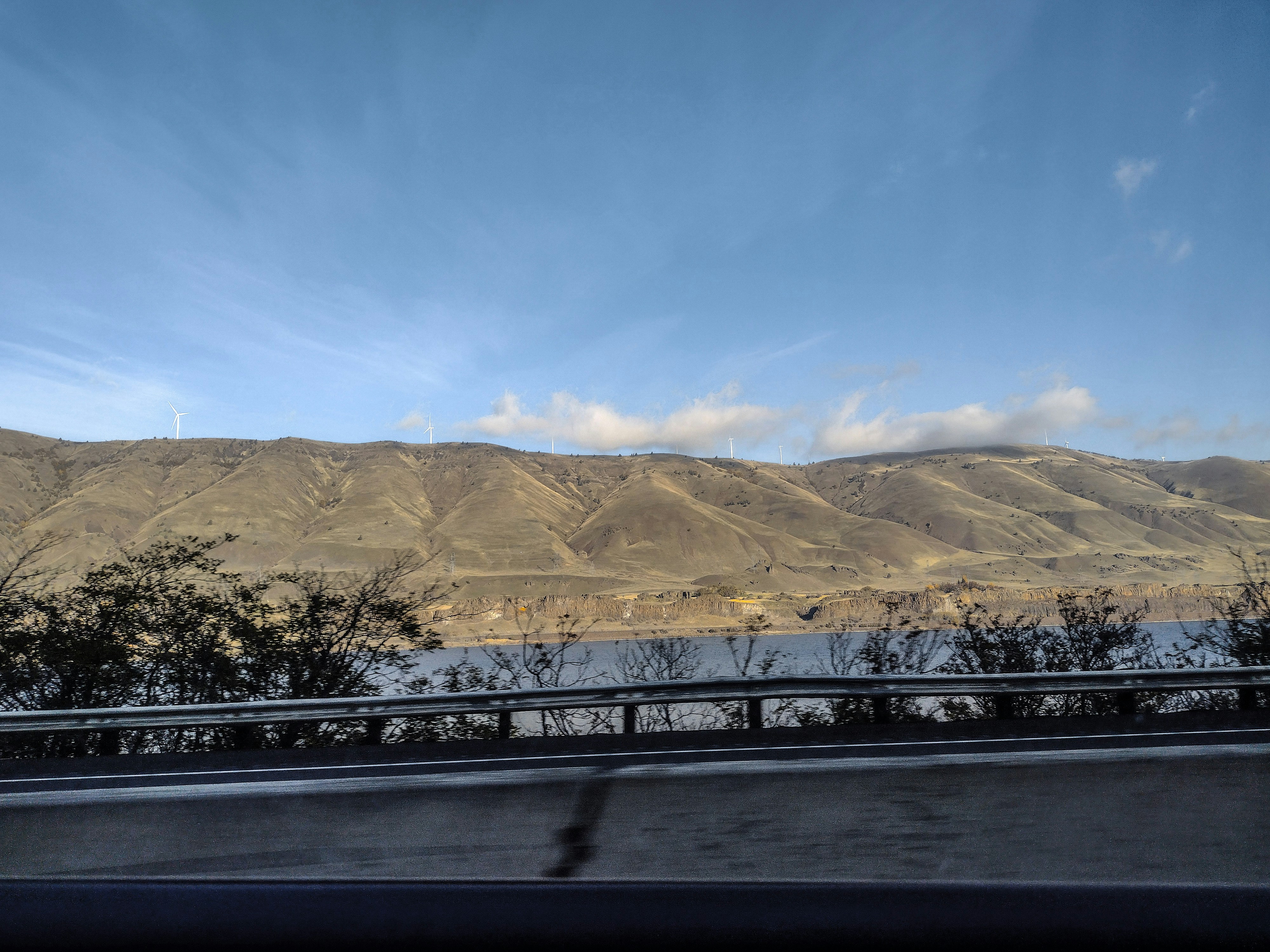 Hills stretch across the horizon dotted with wind turbines, as seen from a roadside guardrail against a bright, clear sky.