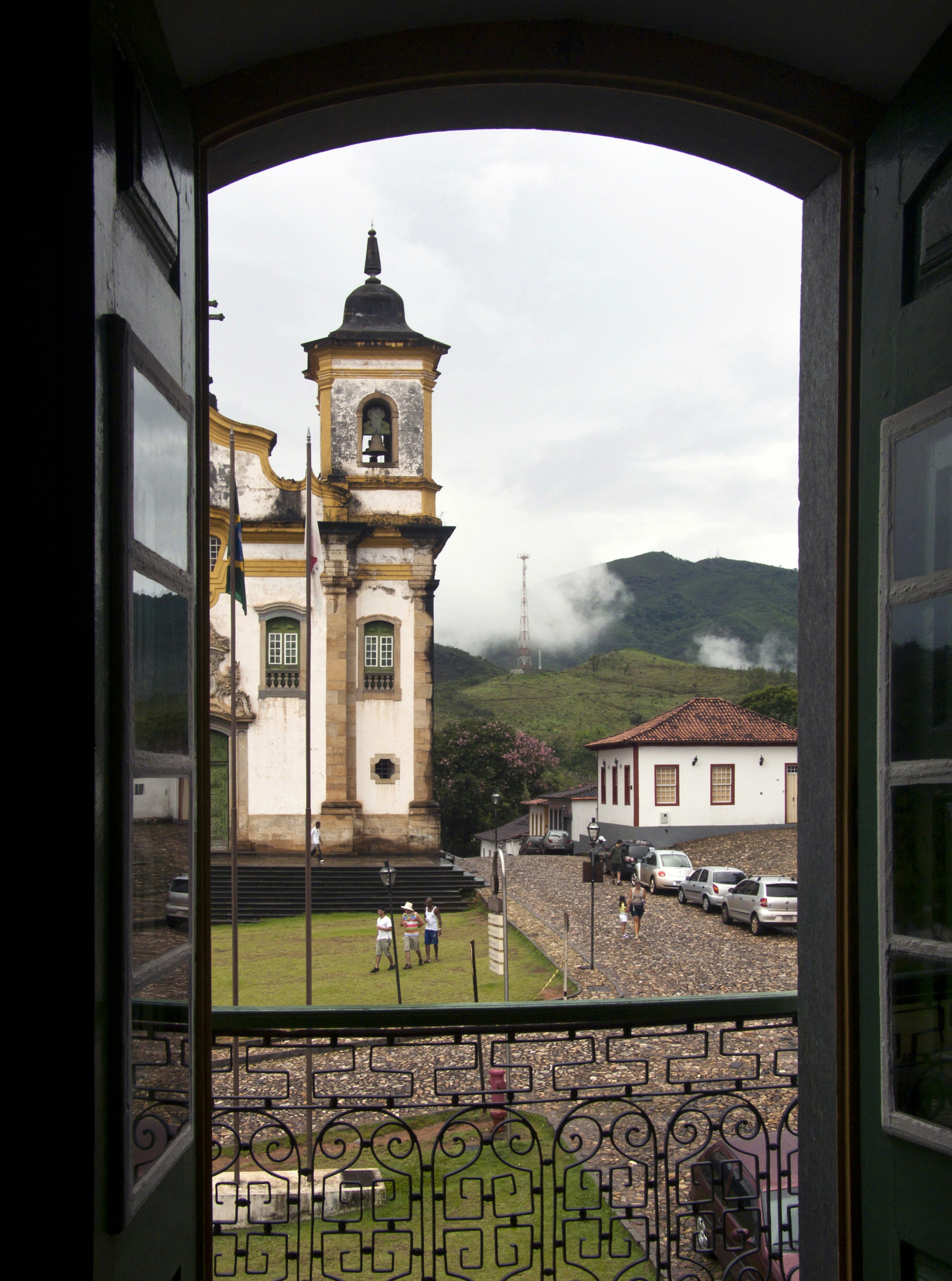Colonial church with a tall bell tower is framed by an open archway and decorative railing. People stroll a cobbled plaza while cars line the street against a hillside and cloudy sky.