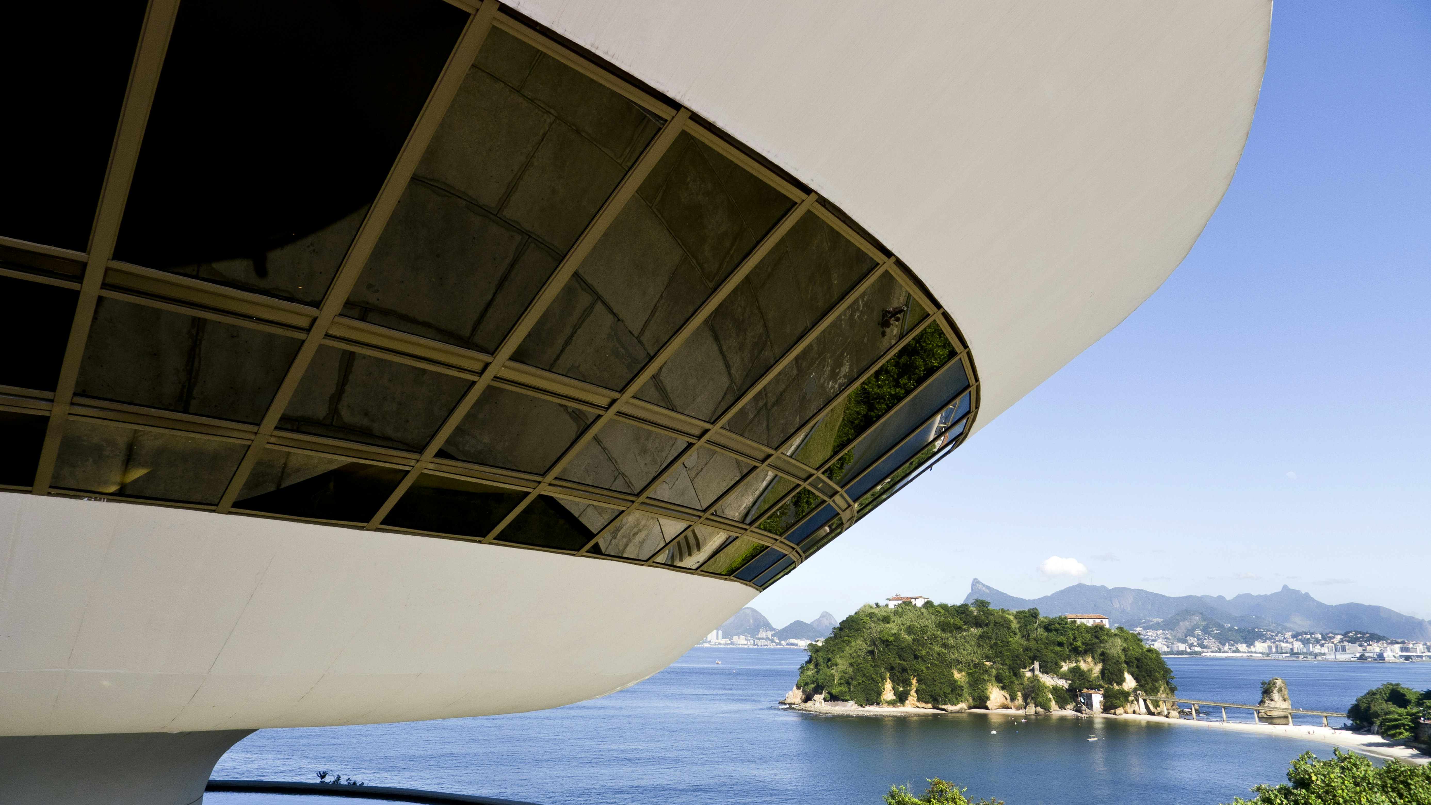 Modern building façade overlooking a tranquil bay with distant mountains under a clear sky.