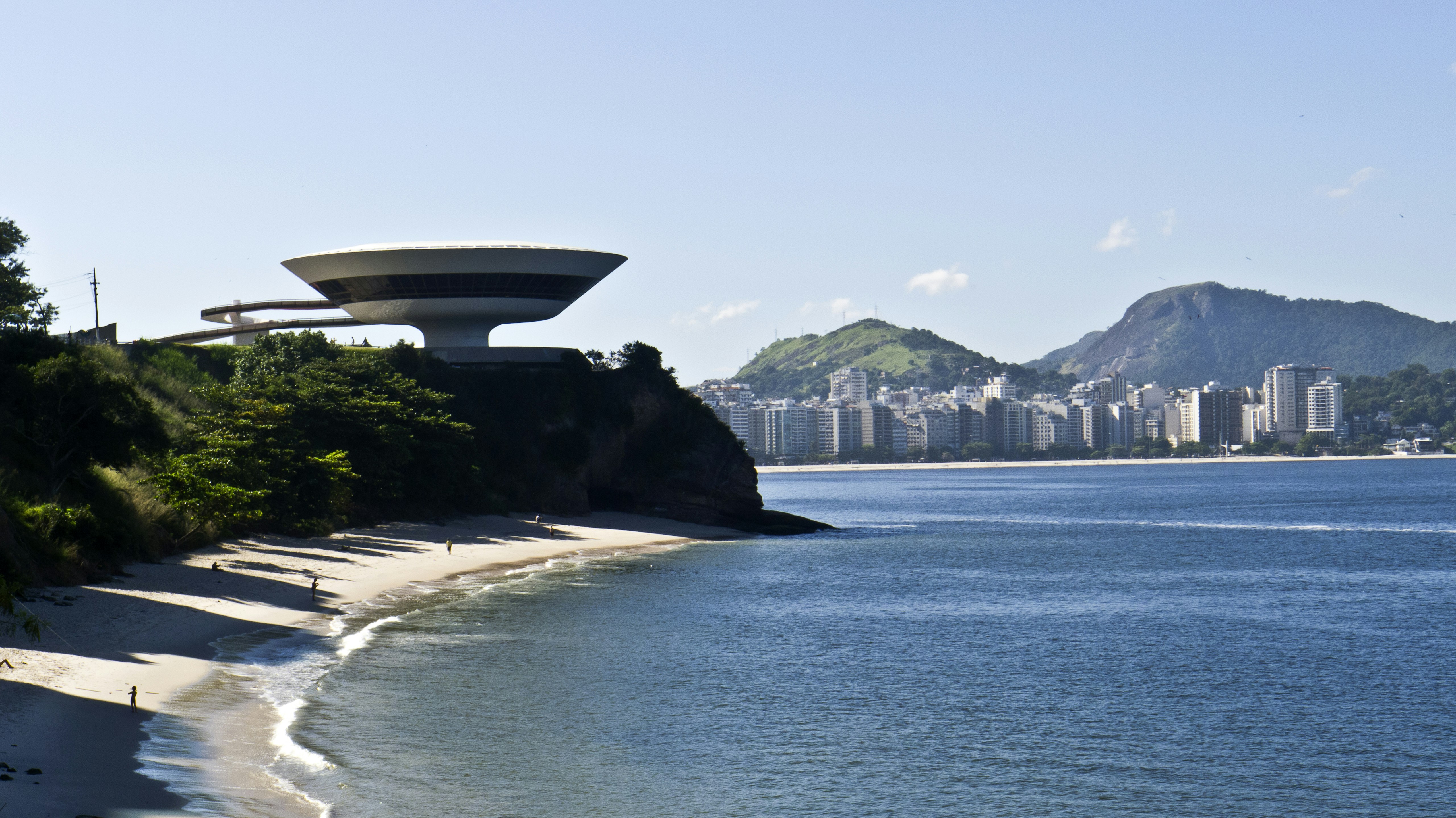 A striking modernist structure perched on a coastal cliff, framed by a serene beach and distant city skyline. The scene captures the harmony between architecture and nature.