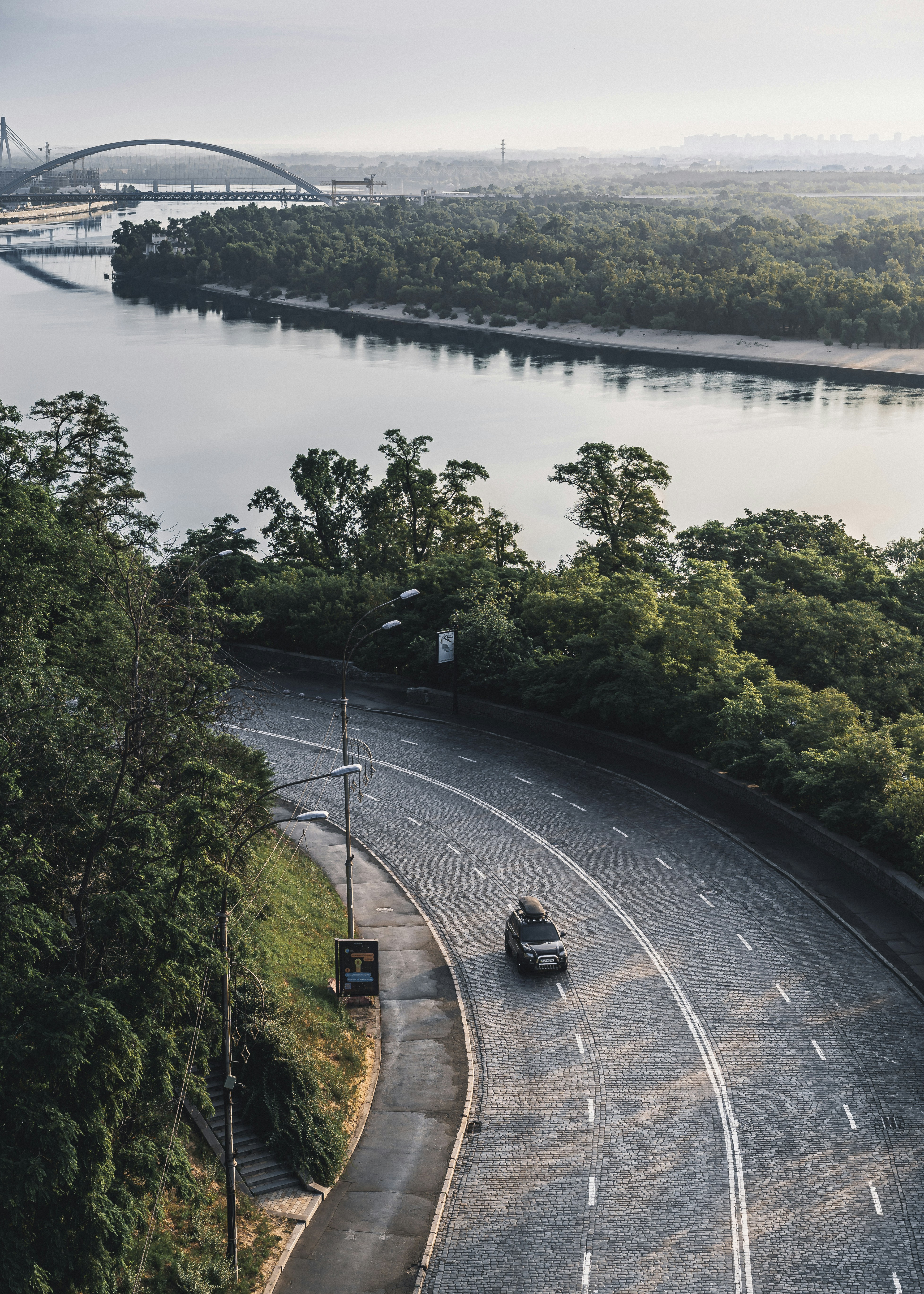 Winding road curves alongside a tranquil river with lush greenery and a distant bridge under soft morning light.