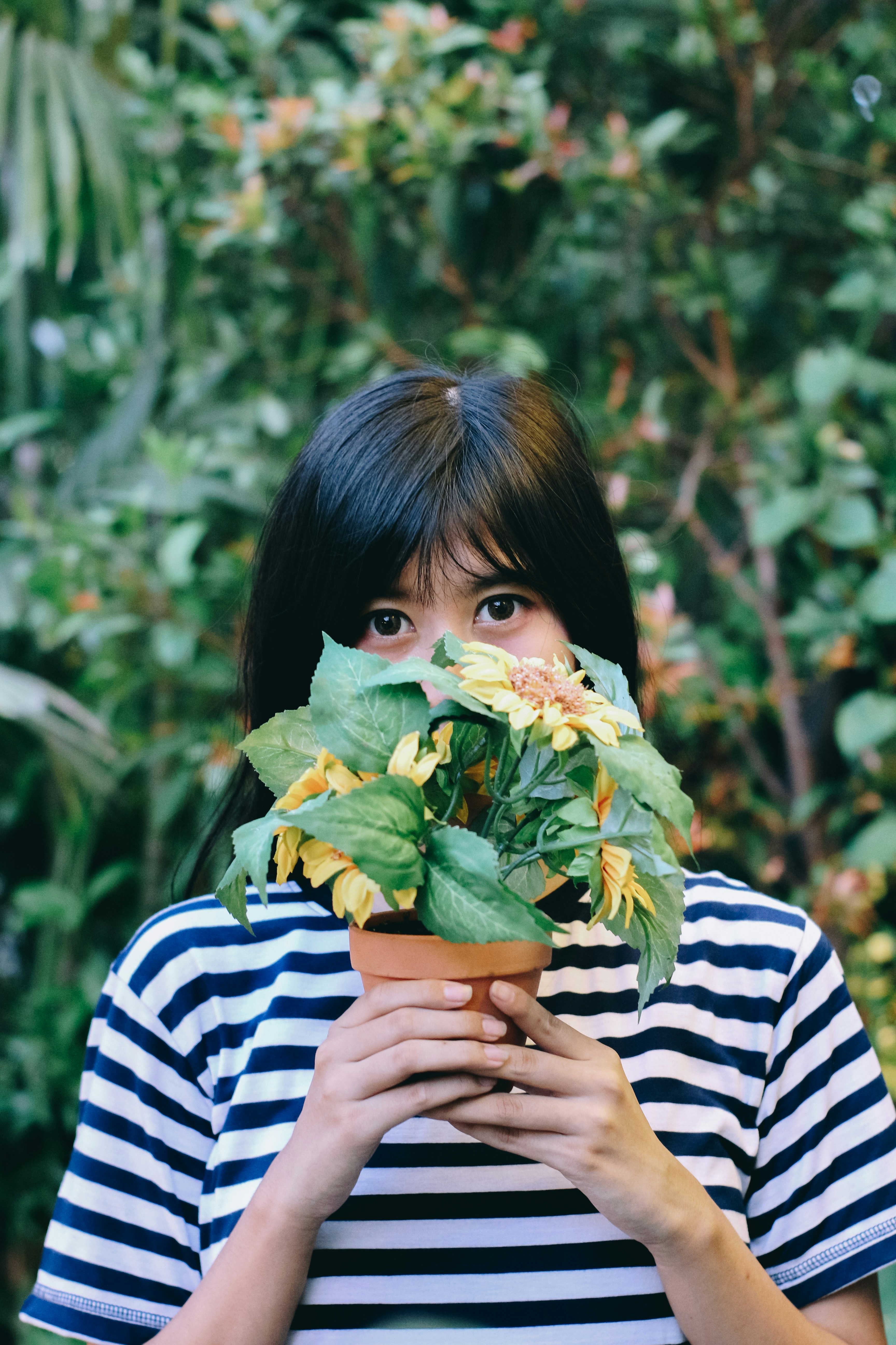 A young woman cradles a potted flower, partially obscured by its vibrant leaves, set against a lush green backdrop.