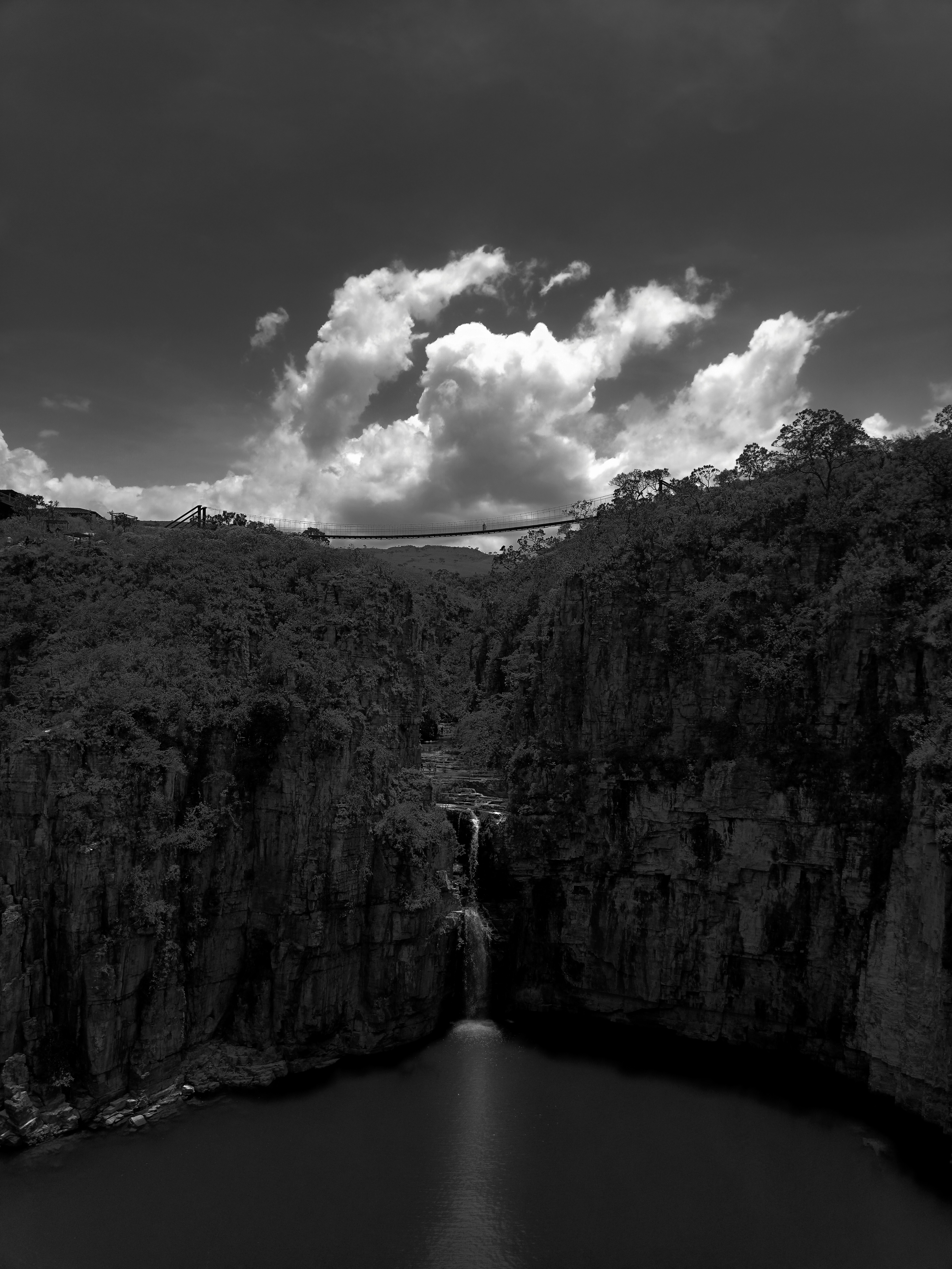 Monochrome scene of a deep canyon with tall rock walls and a narrow river at the bottom, a bridge visible near the horizon and dramatic clouds overhead.