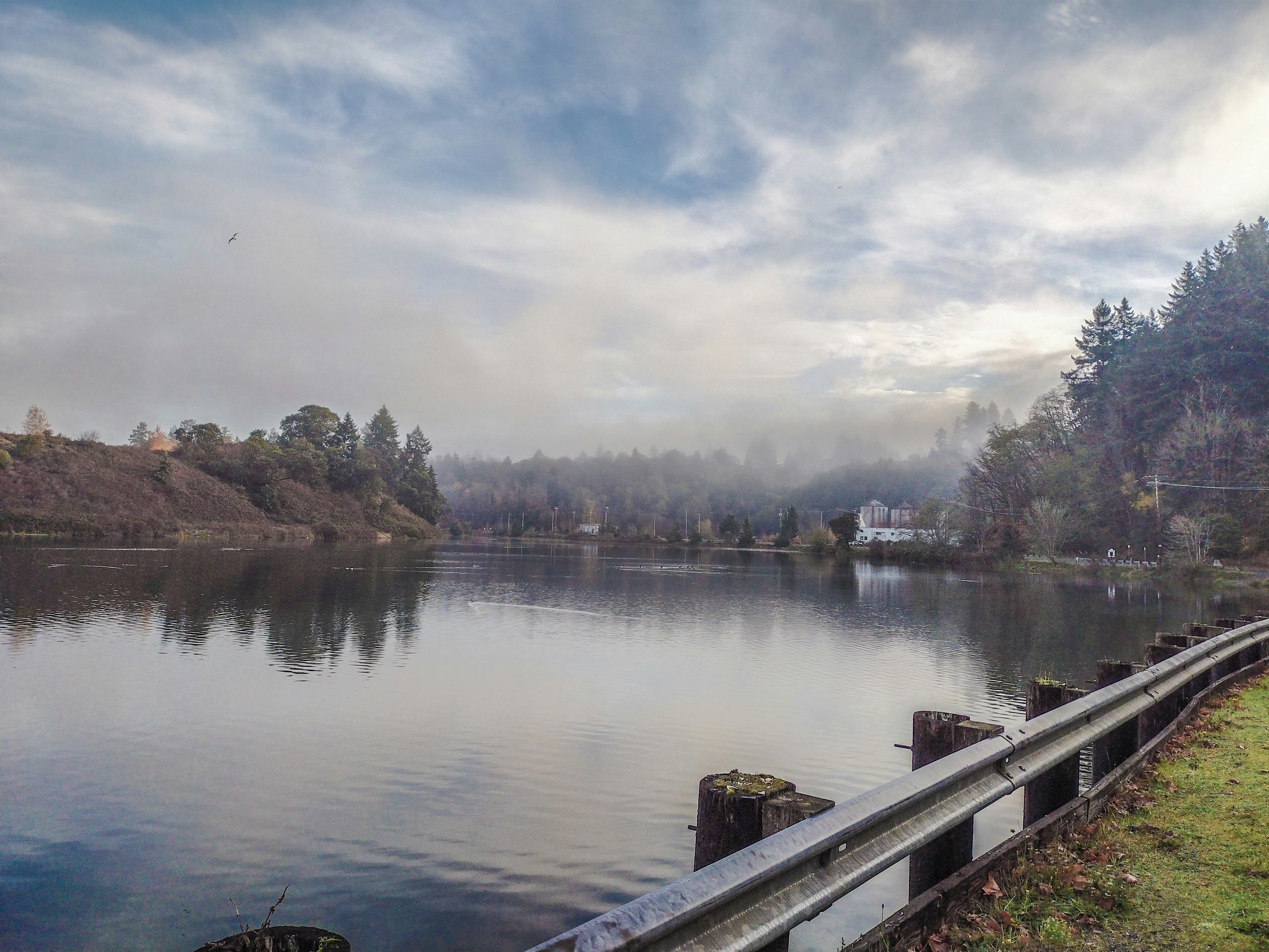 A tranquil lakeside photograph taken from a railing along the water, with misty hills and a distant white house reflected on the calm lake.