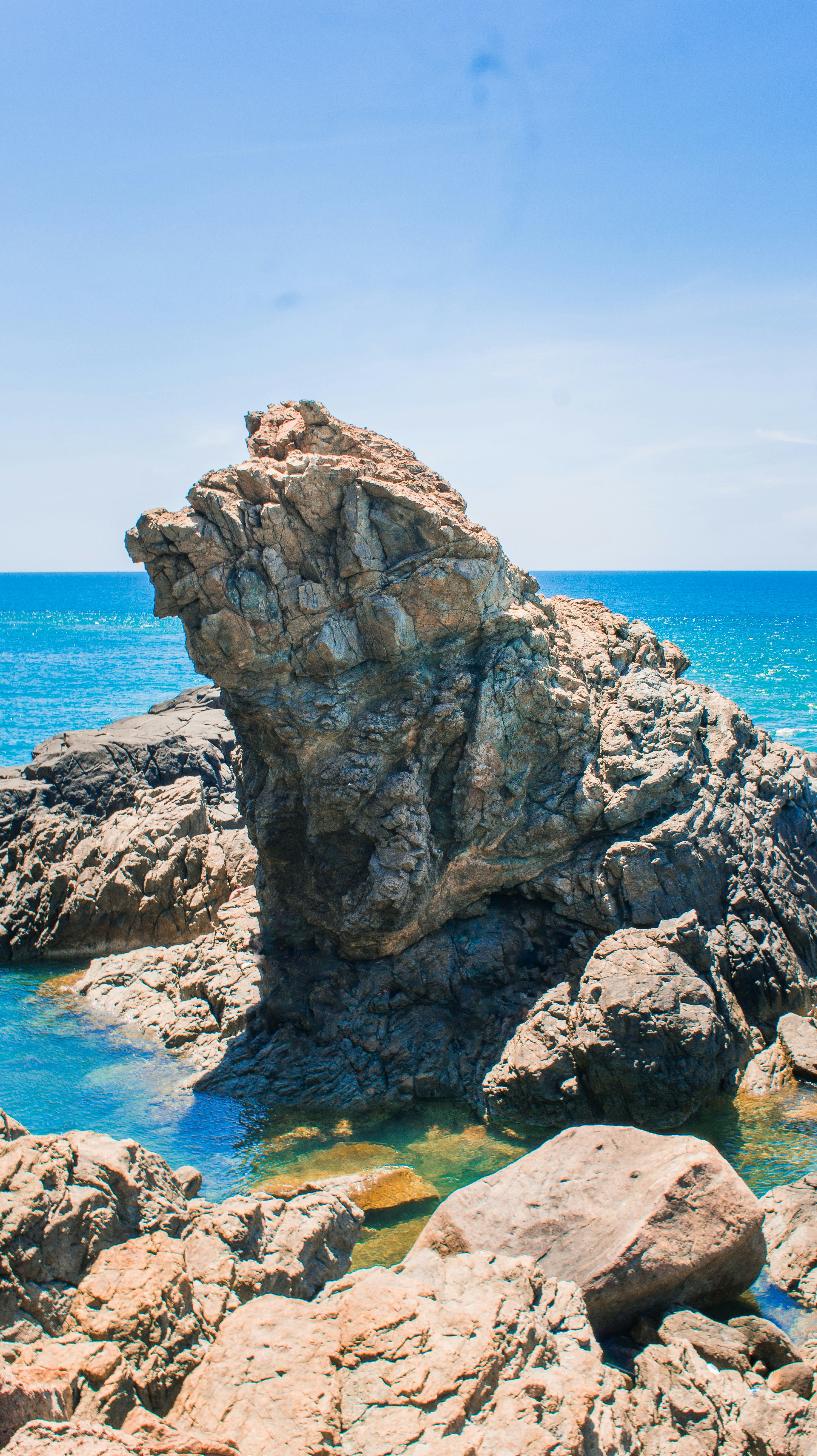 A rugged rock formation stands sentinel over the tranquil waters, surrounded by a mosaic of stones and clear blue sea.