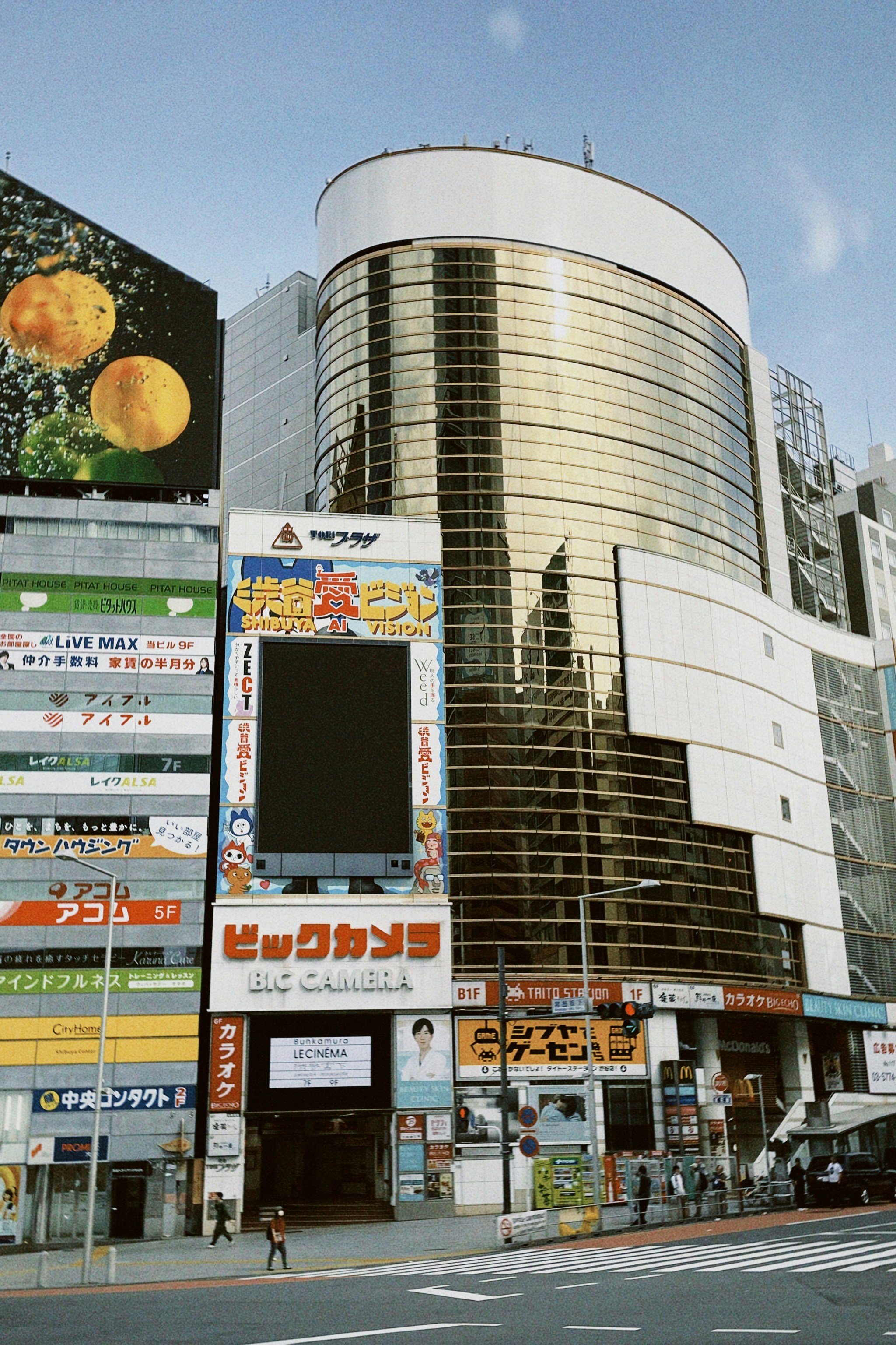 Photo of a curved glass building with large neon billboards and a busy street scene.