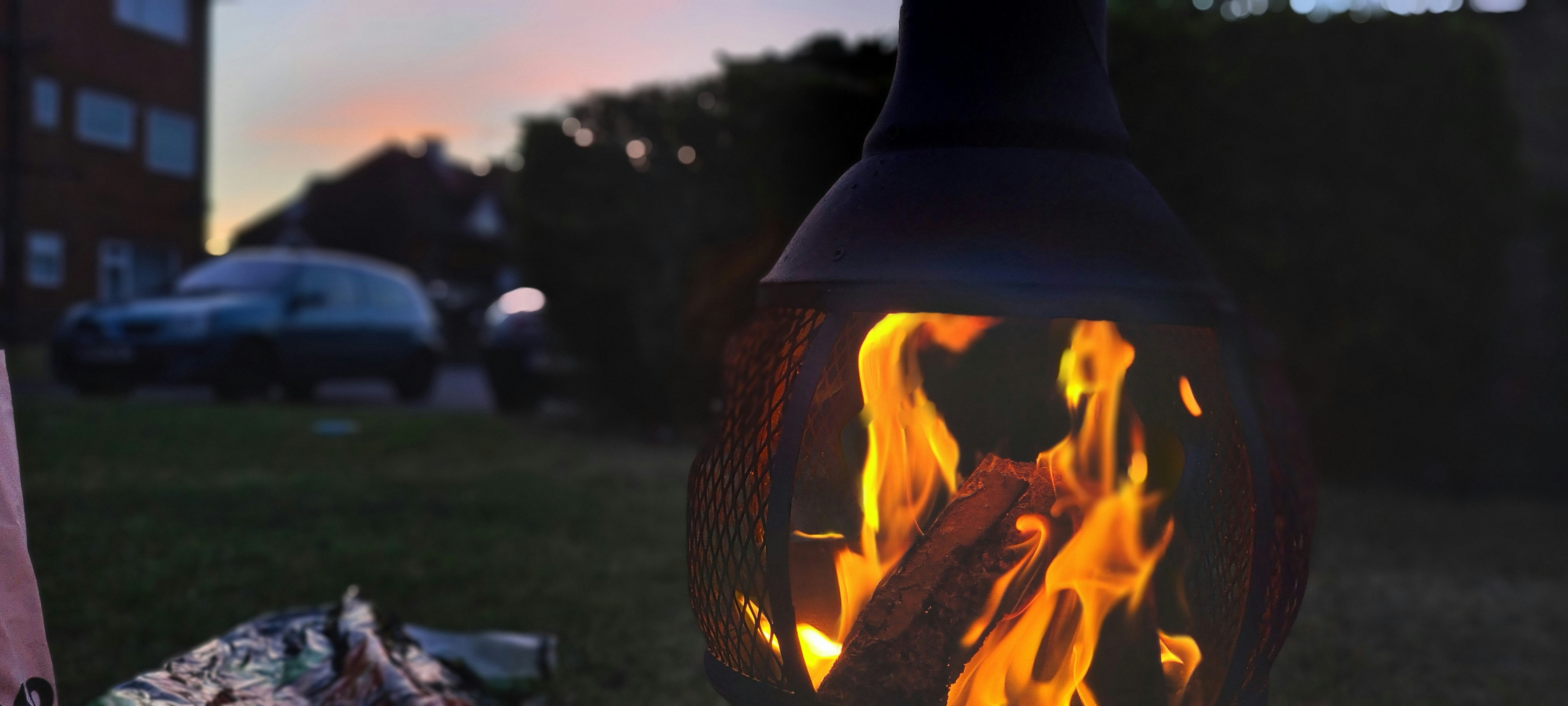 Flames dance inside a chiminea as the sun sets, with silhouetted trees and buildings in the background.