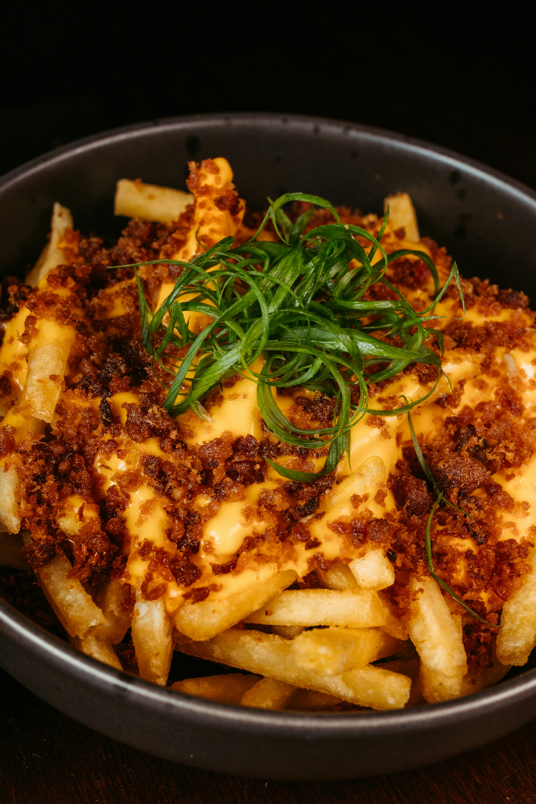 Crumbled Bacon Cheesed Fries with Green Garnish in a Black Bowl