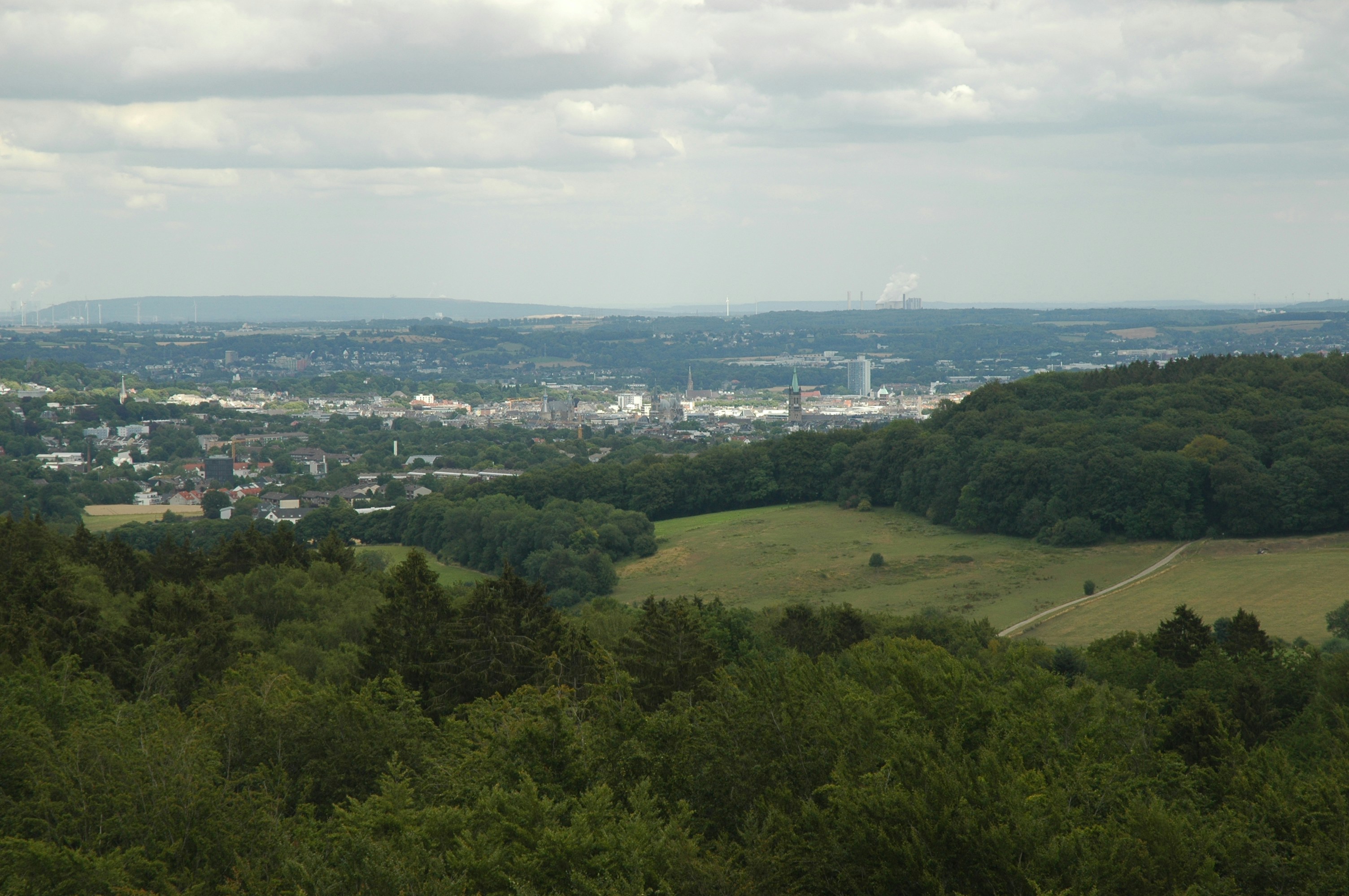 Cityscape viewed from a hill, with lush greenery in the foreground and scattered clouds above.
