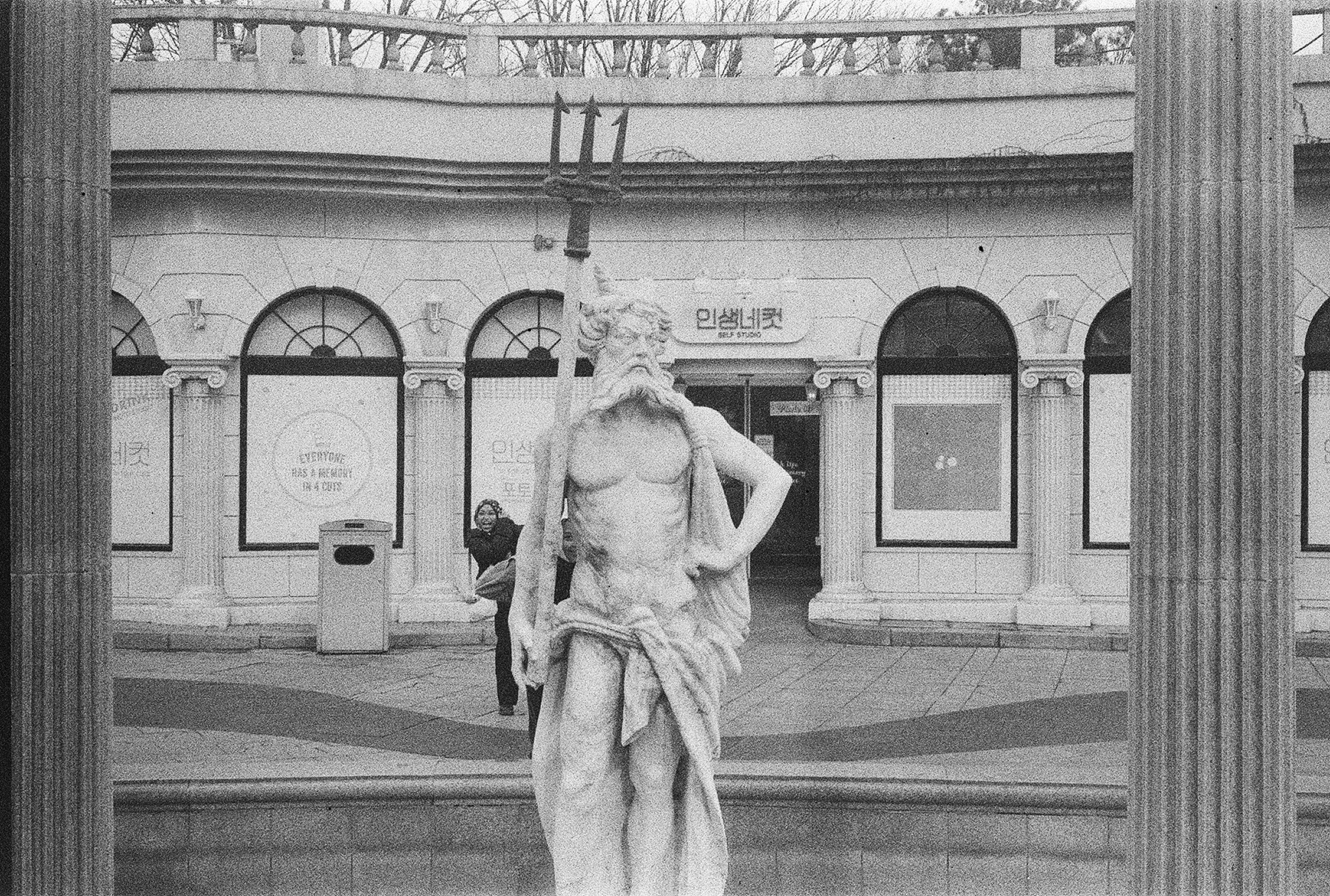 Black-and-white photograph of a classical statue centered among arched storefronts and columns in a quiet urban plaza. The composition emphasizes the sculpture against the architectural backdrop.