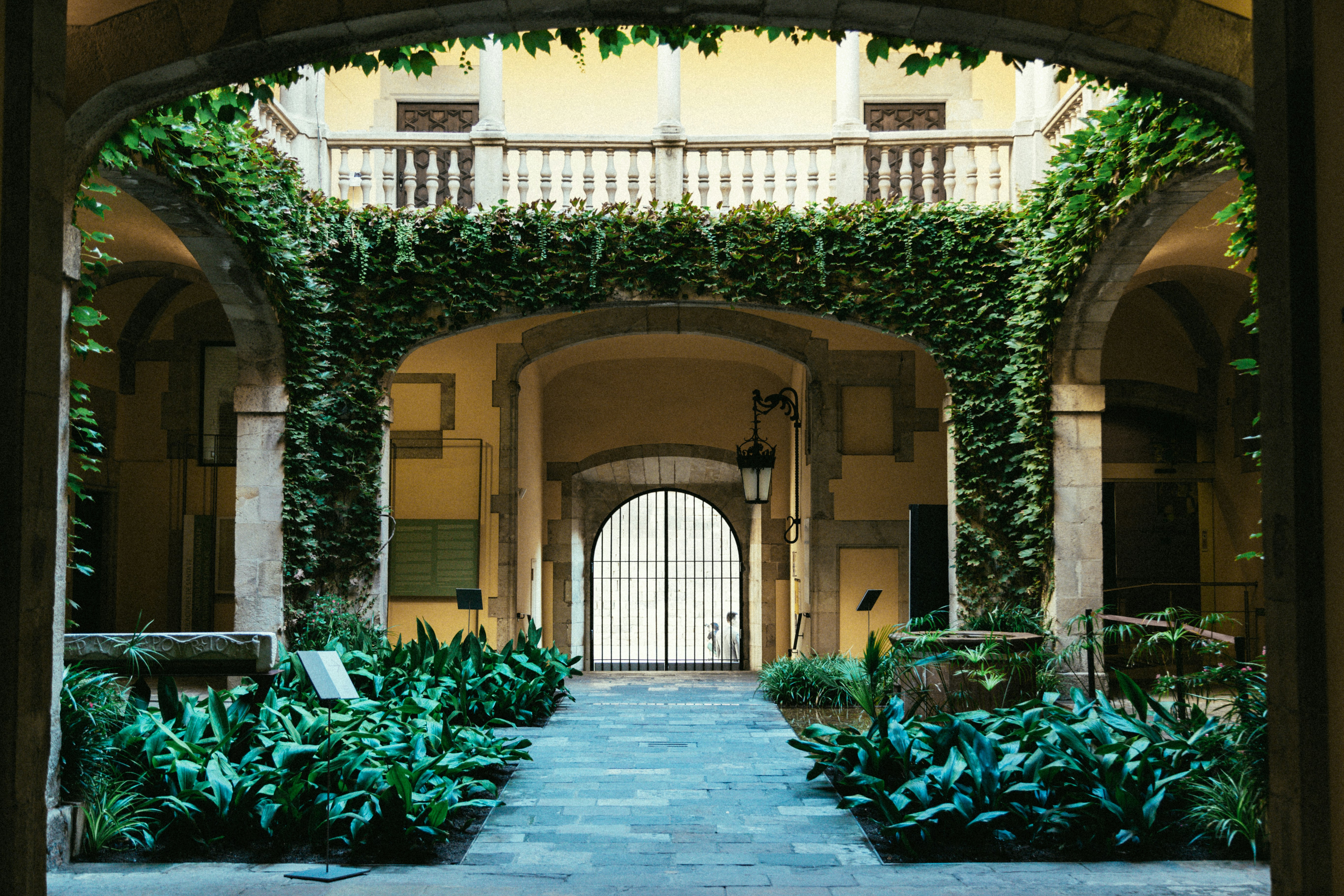 Lush greenery frames a serene courtyard entrance, leading to a gated passageway. The architecture blends nature and structure harmoniously.