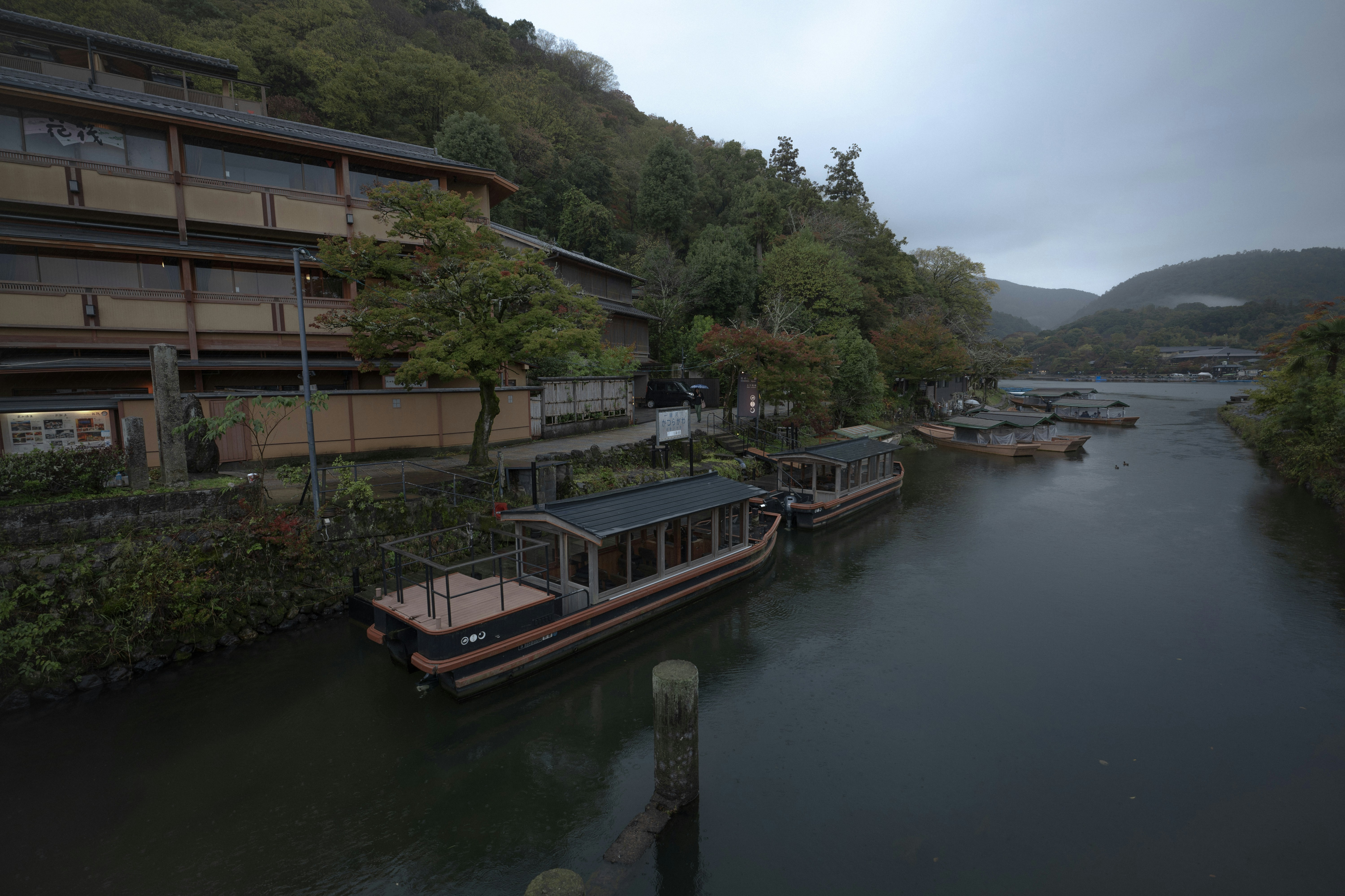 A riverside scene with traditional moored boats along a calm canal, flanked by a wooden riverside building and trees under an overcast sky.